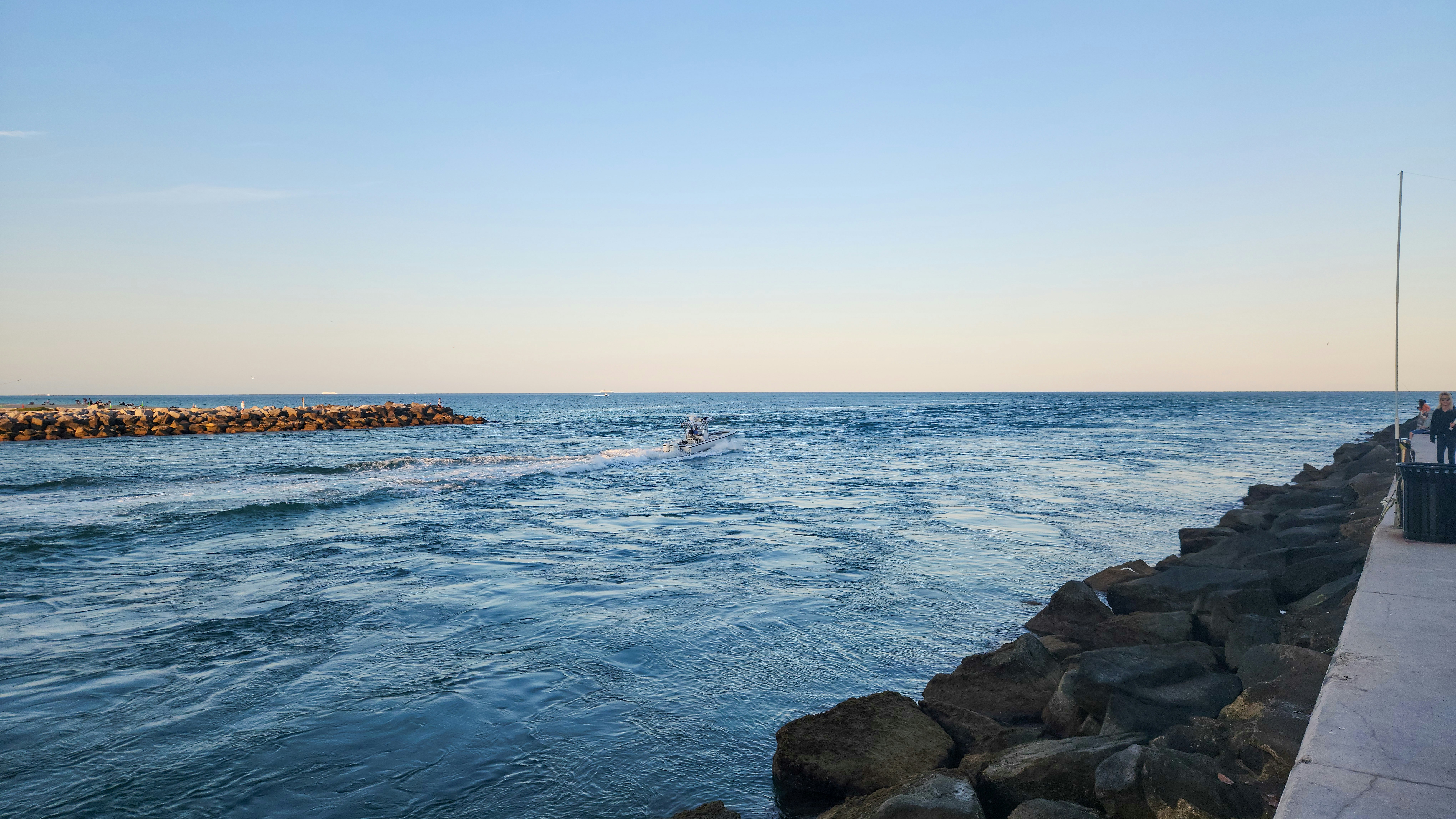 Person standing on a pier with rocky shoreline, overlooking calm sea under a clear sky.