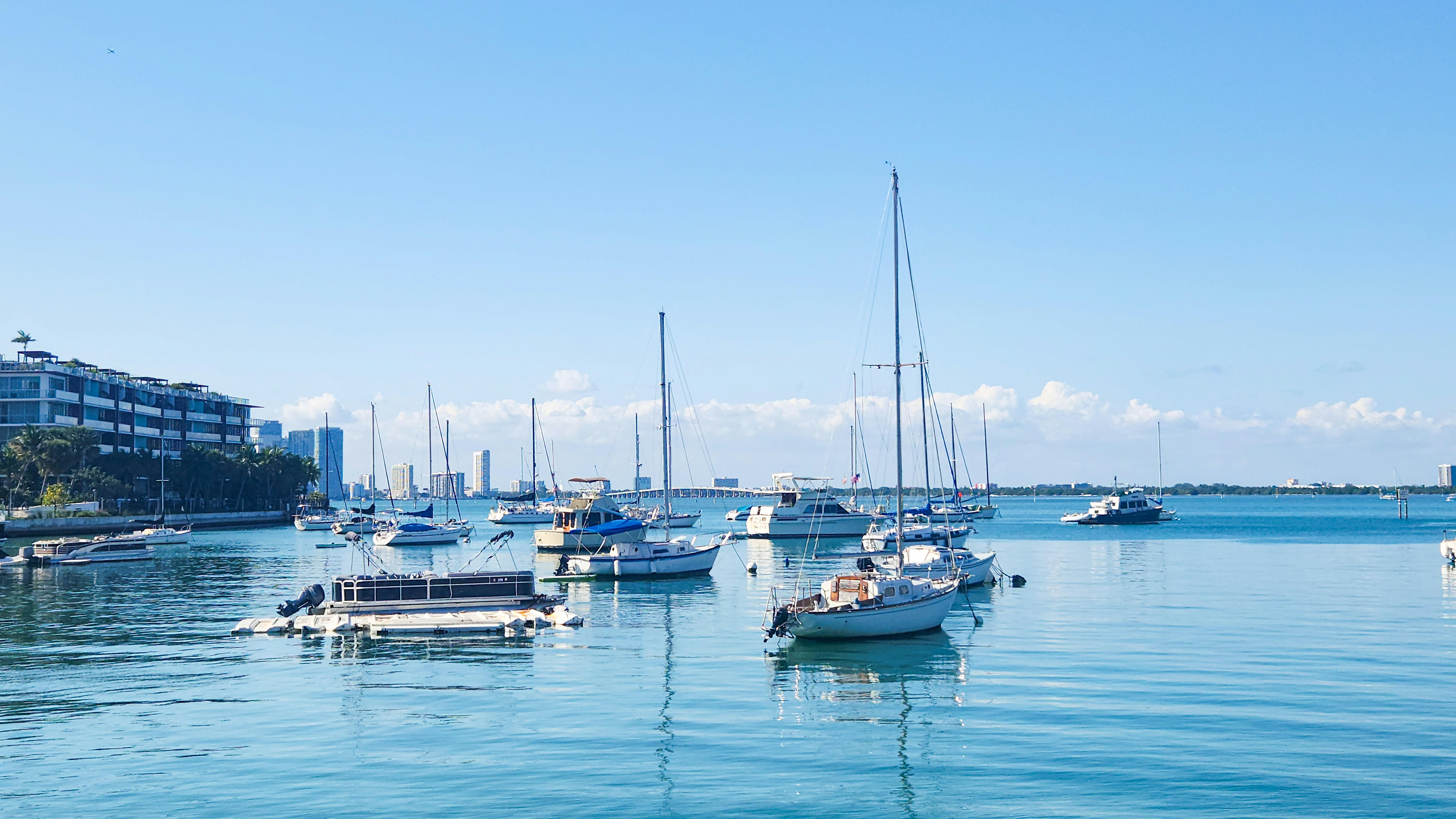 Boats and yachts peacefully anchored in Biscayne Bay with Miami skyline in the background under a clear blue sky.