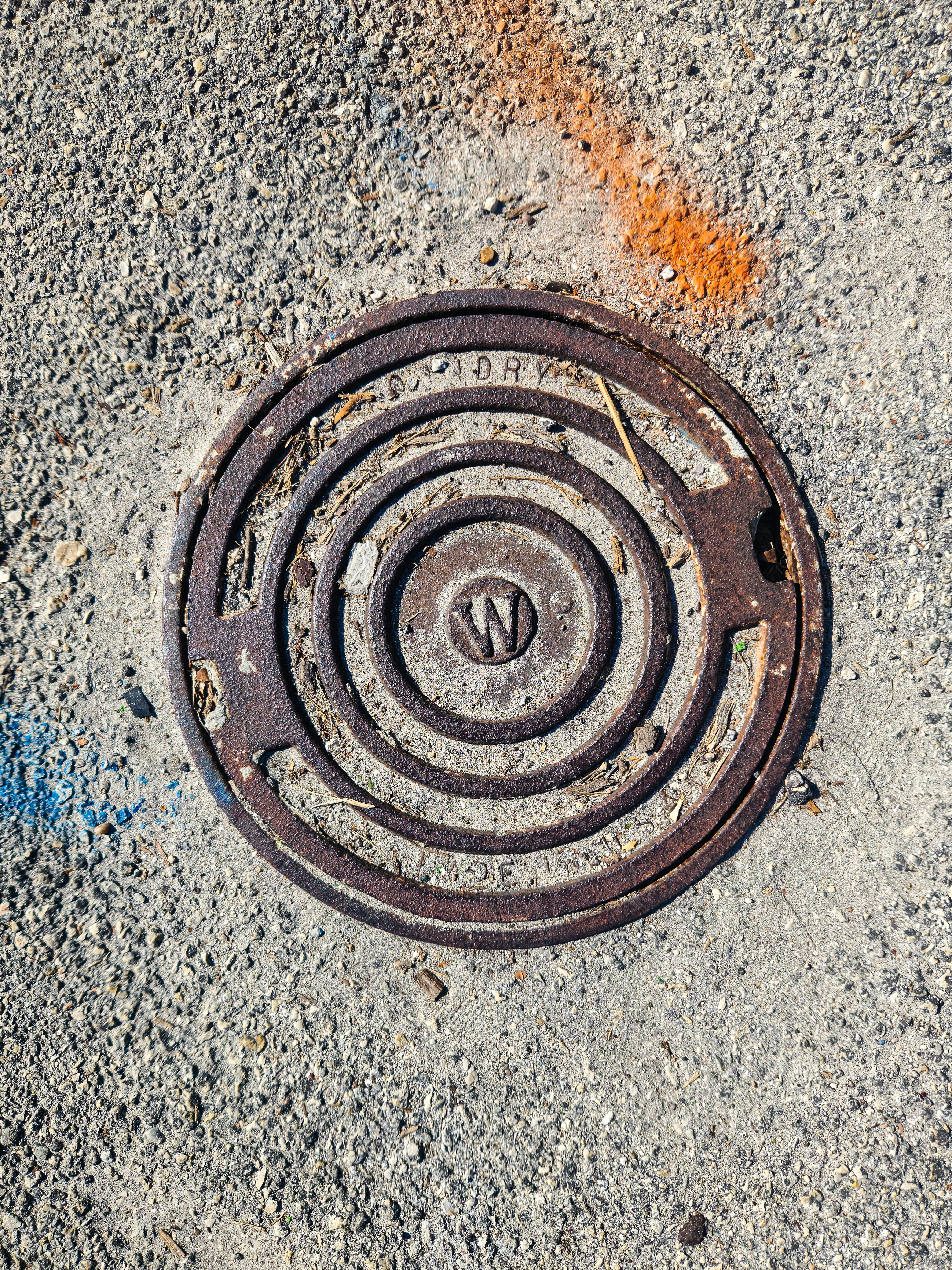 Rusty manhole cover with concentric circles and a central emblem, set against a textured concrete surface with splashes of color.