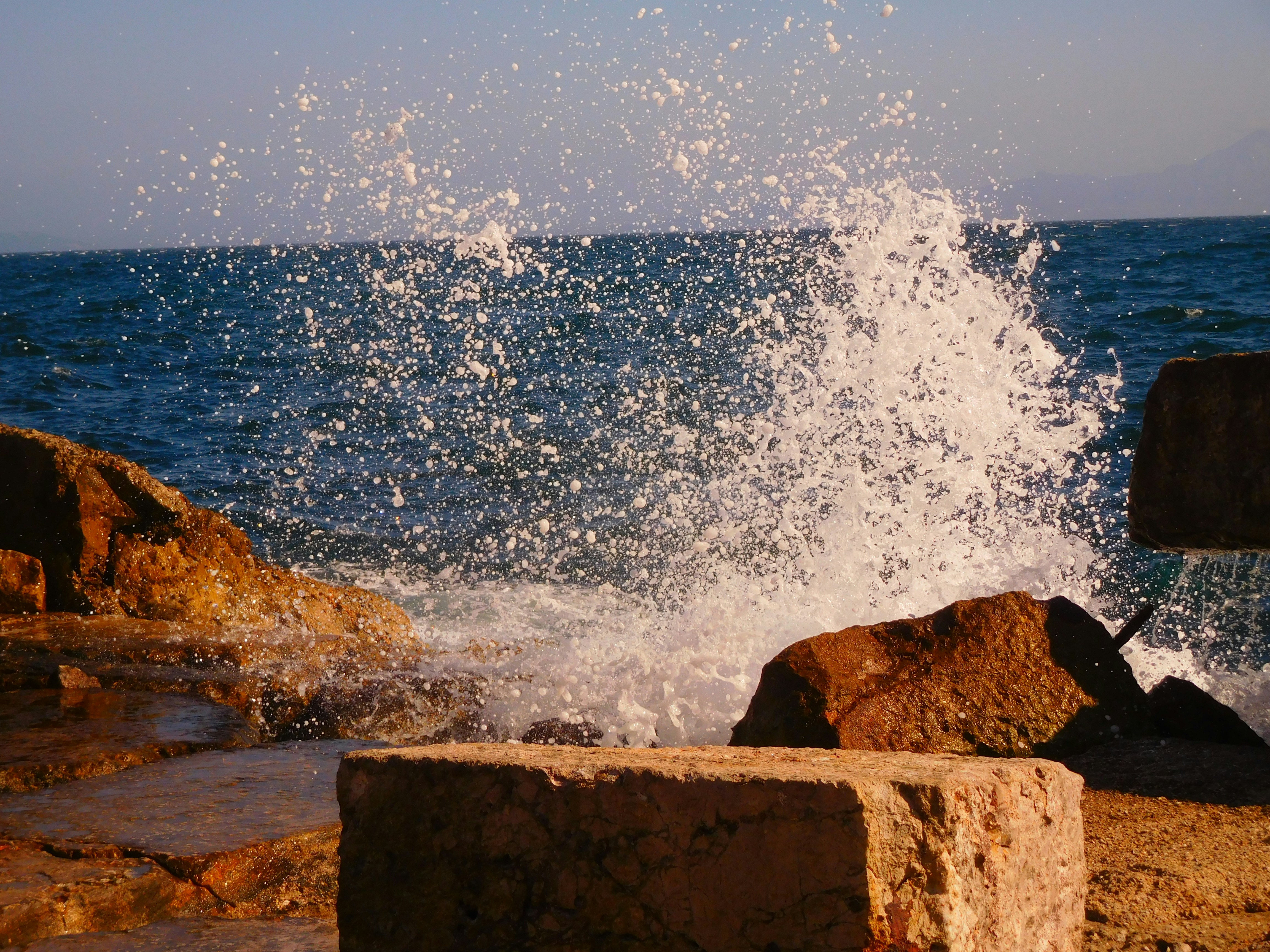 Waves crashing against rugged rocks with water droplets suspended mid-air in bright sunlight.