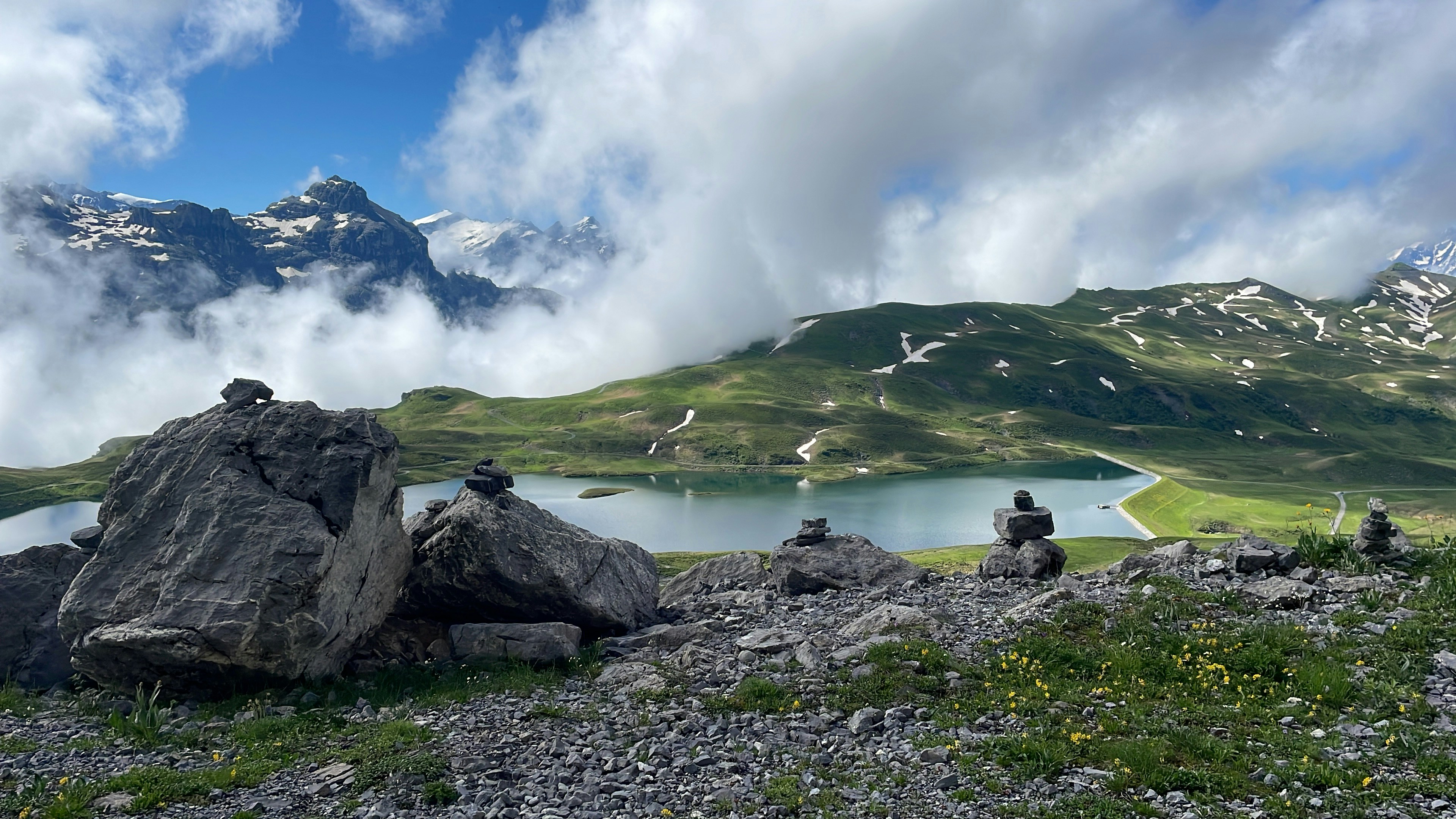 Rock formations overlook a tranquil lake surrounded by snow-speckled mountains beneath a cloud-strewn sky.