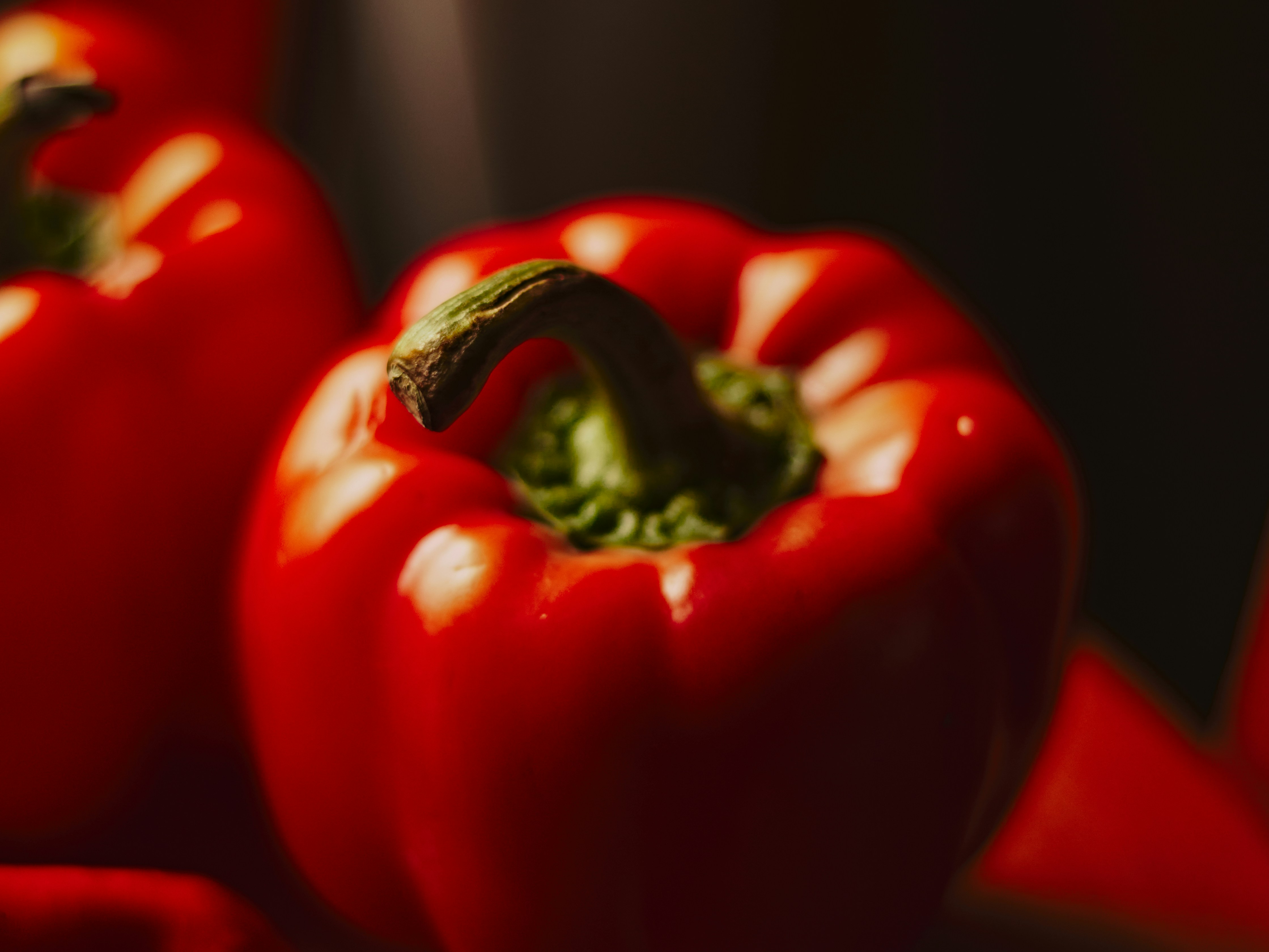 Two red bell peppers on a textured surface, with one in sharp focus and the other slightly blurred, under warm lighting that highlights their contours.