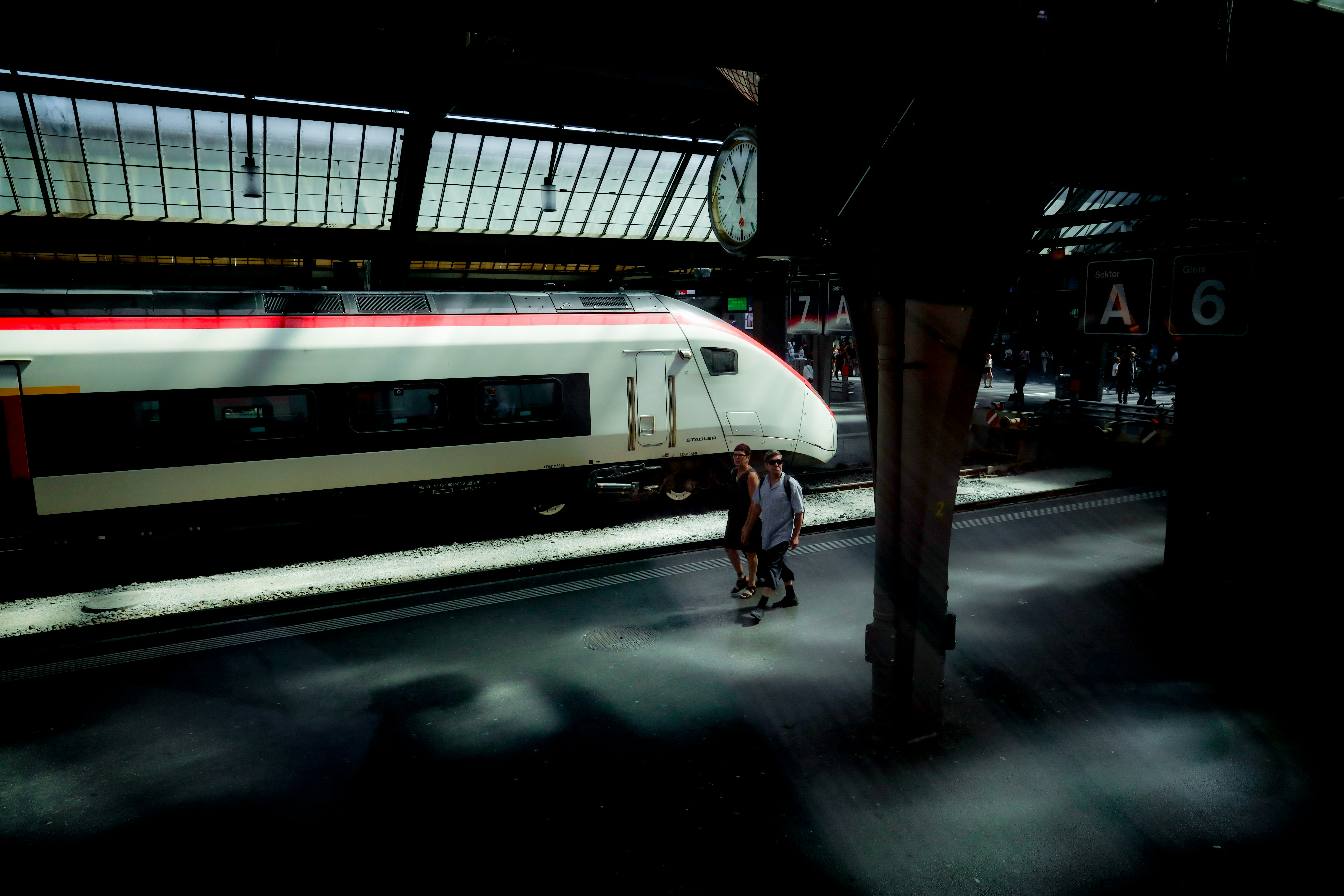 Train station scene with a sleek white train under a glass roof, casting dramatic shadows on the platform.