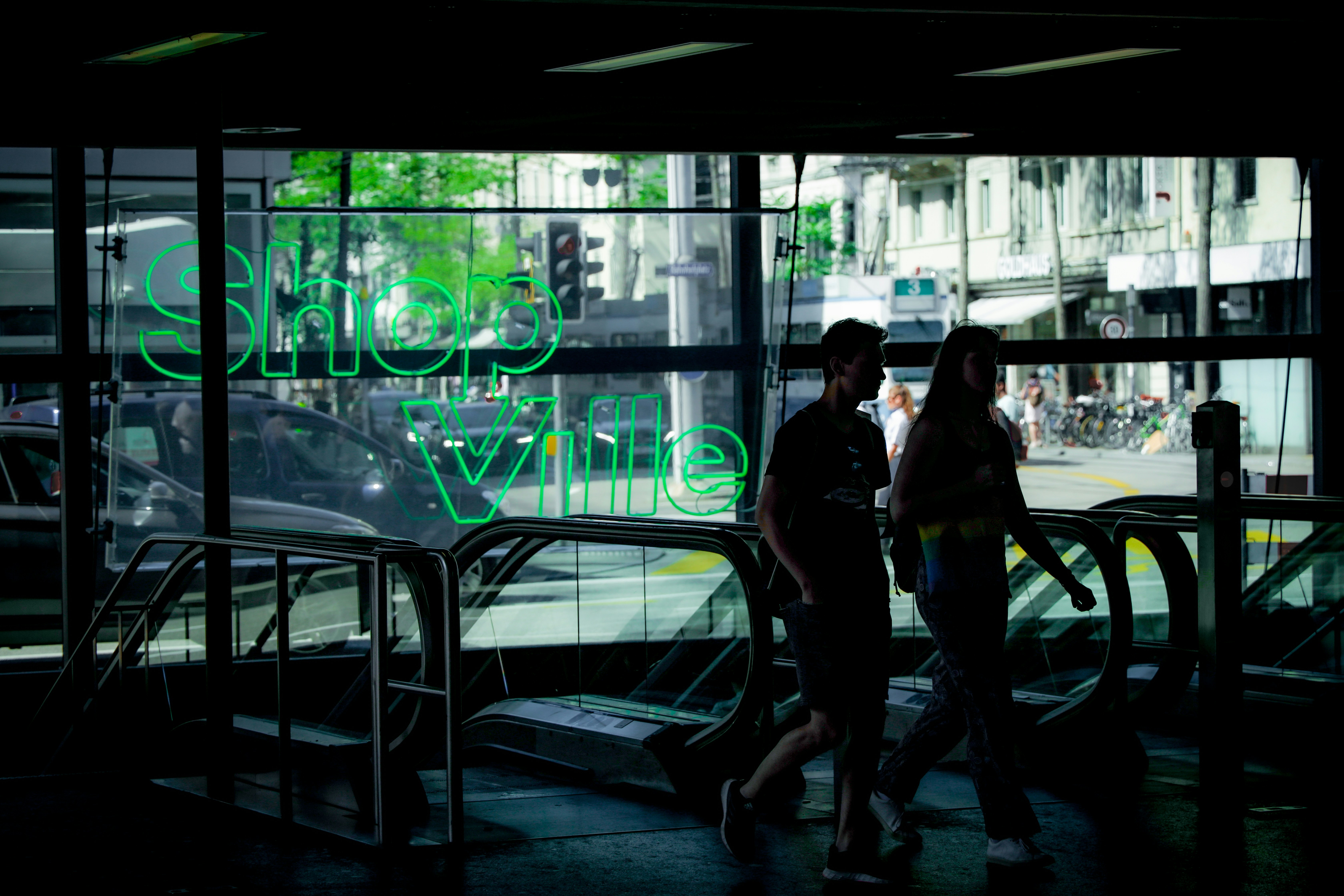 Silhouetted figures walk past neon green signage in a bustling city transit area.
