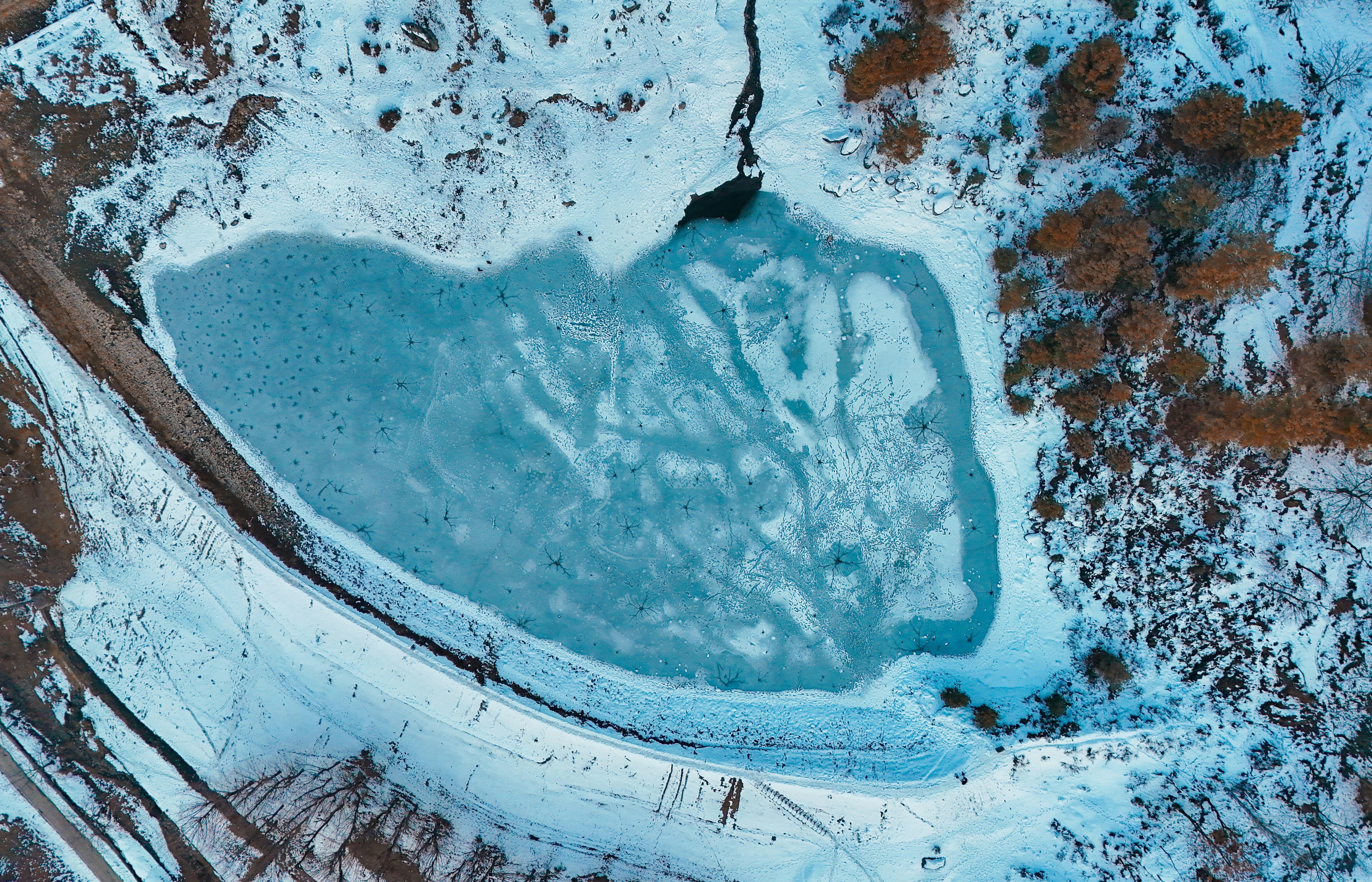 Aerial view of a partially frozen lake surrounded by snow and sparse brown foliage.