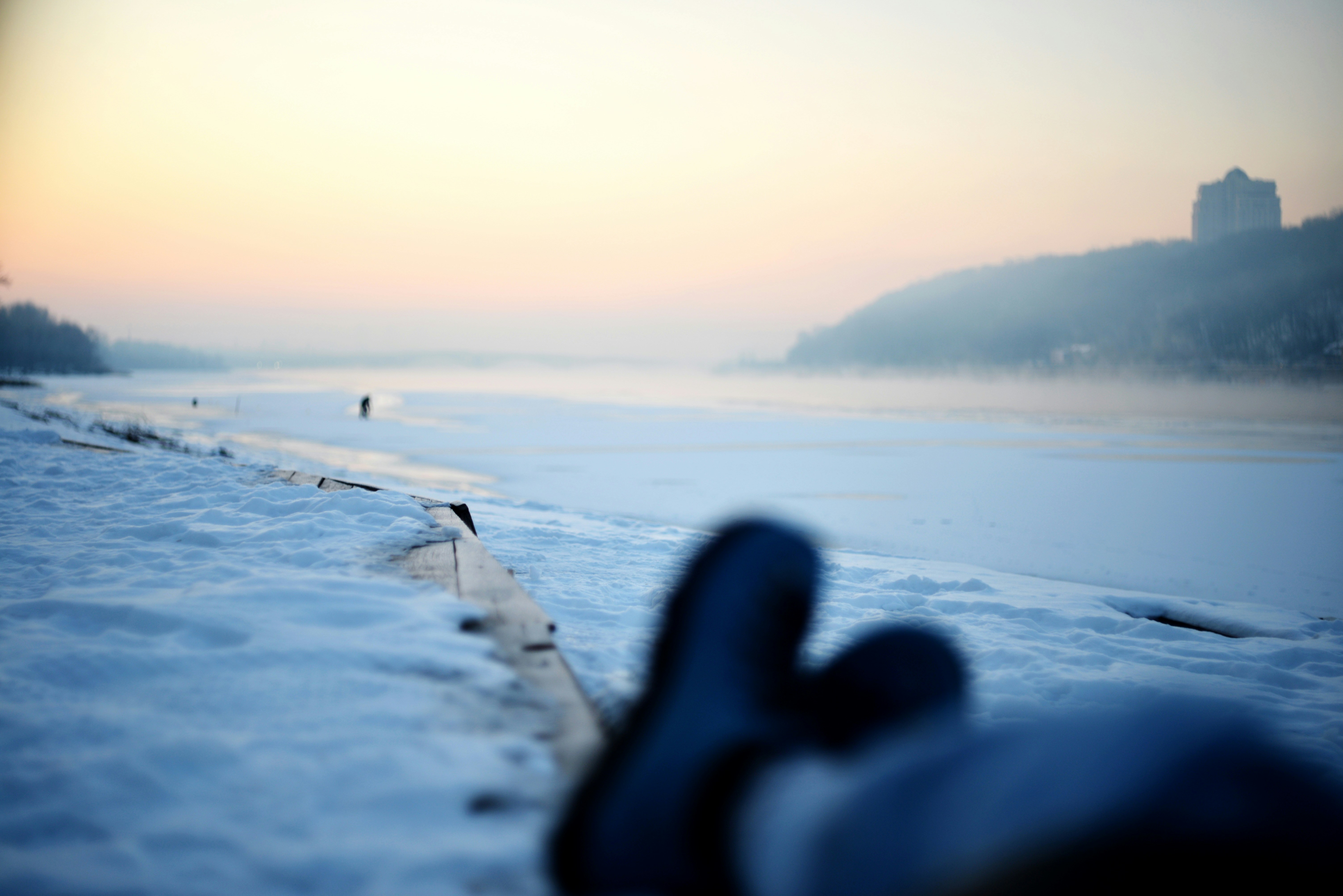 Snowy riverbank at sunrise with soft hues and a distant figure skating on ice.