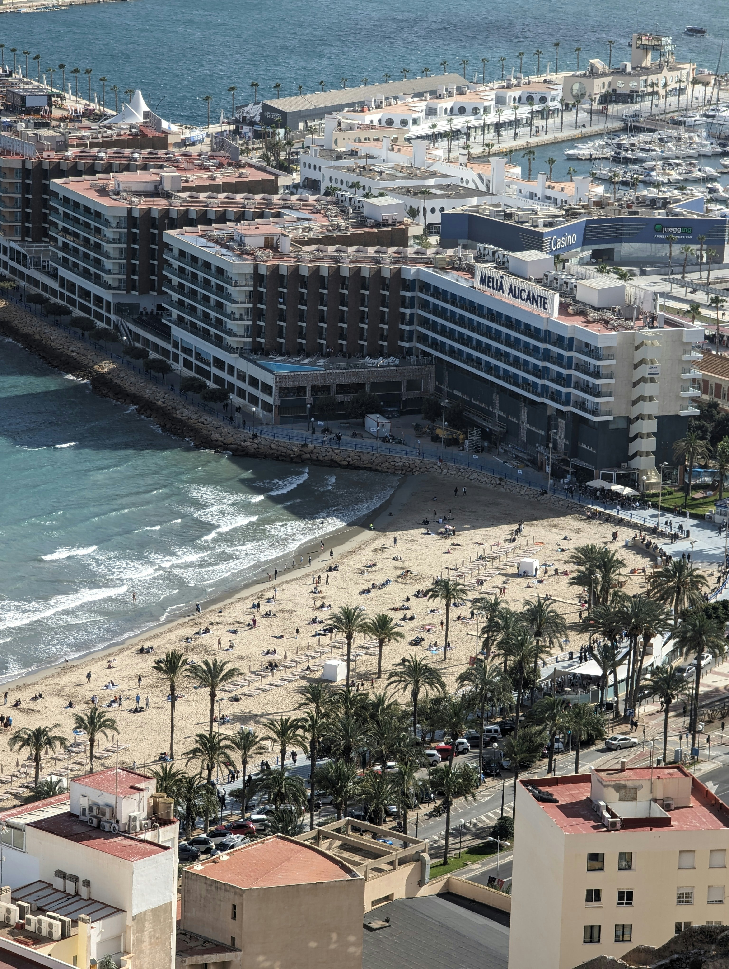 Aerial view of a hotel complex along a sandy beach with palm trees and a distant marina. Coastal resort architecture and a palm-lined promenade frame the shoreline.