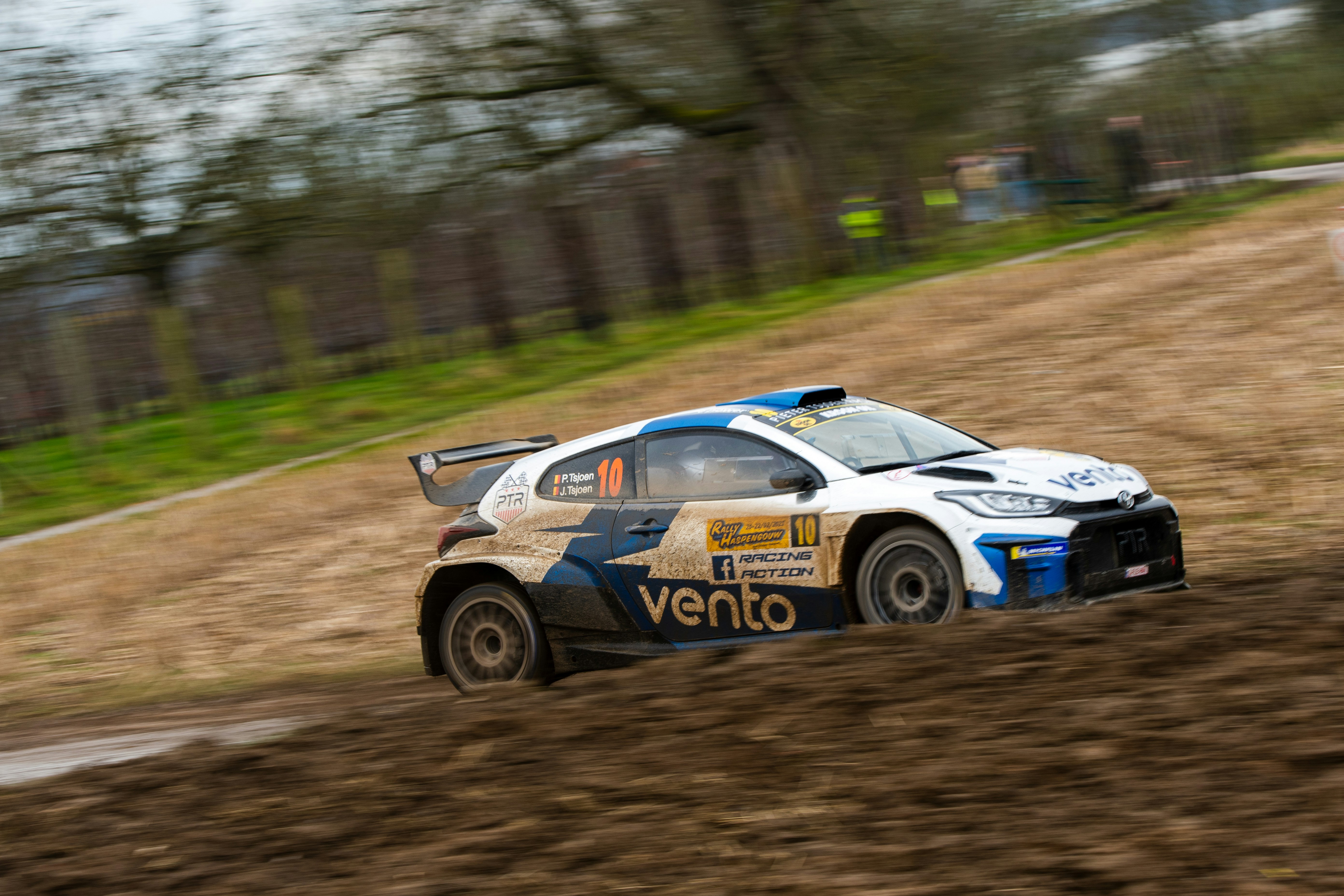 Rally car speeding through a muddy track with blurred motion and trees in the background.