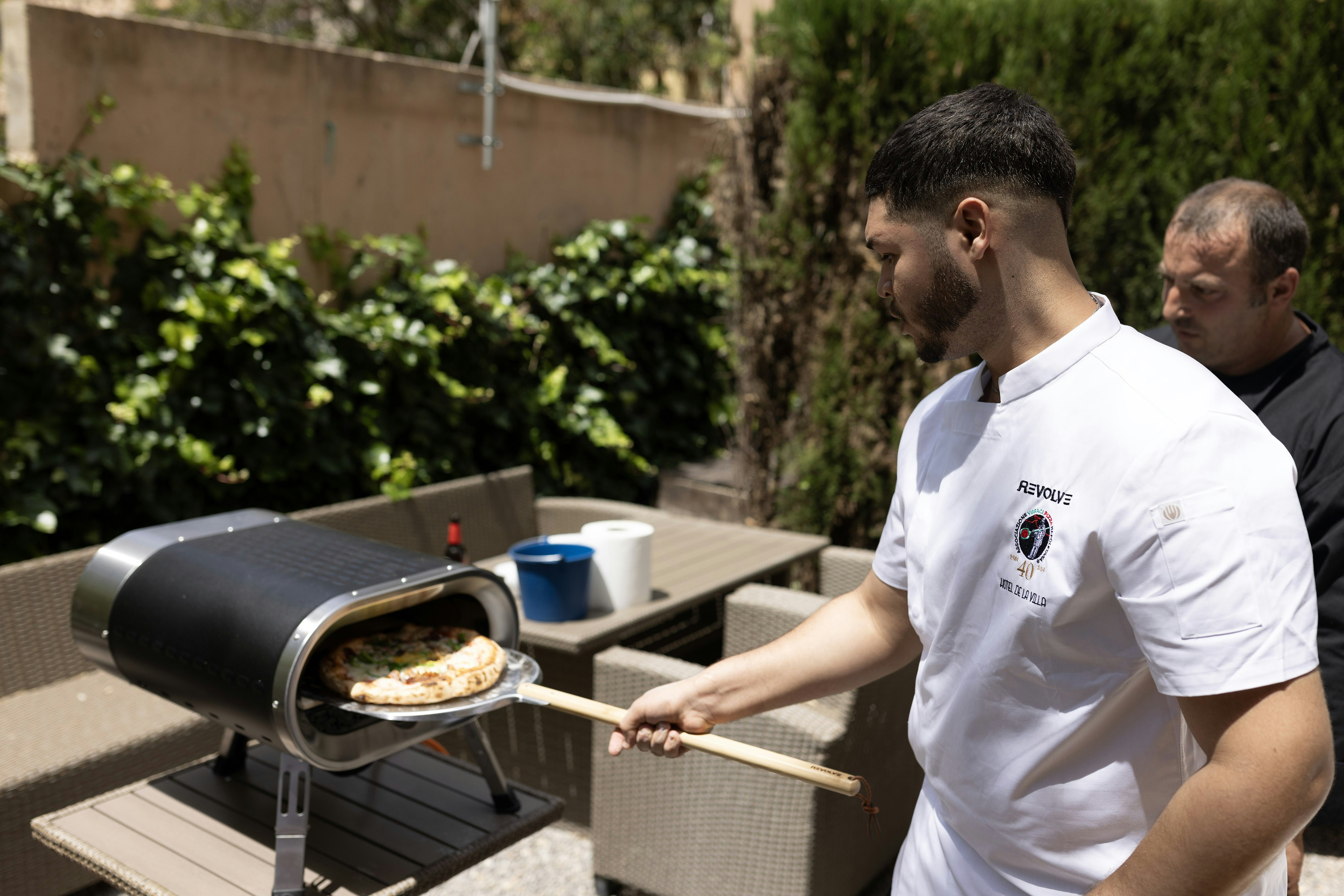 Chef skillfully retrieves a freshly made pizza from a compact outdoor oven, surrounded by lush greenery and modern patio furniture.