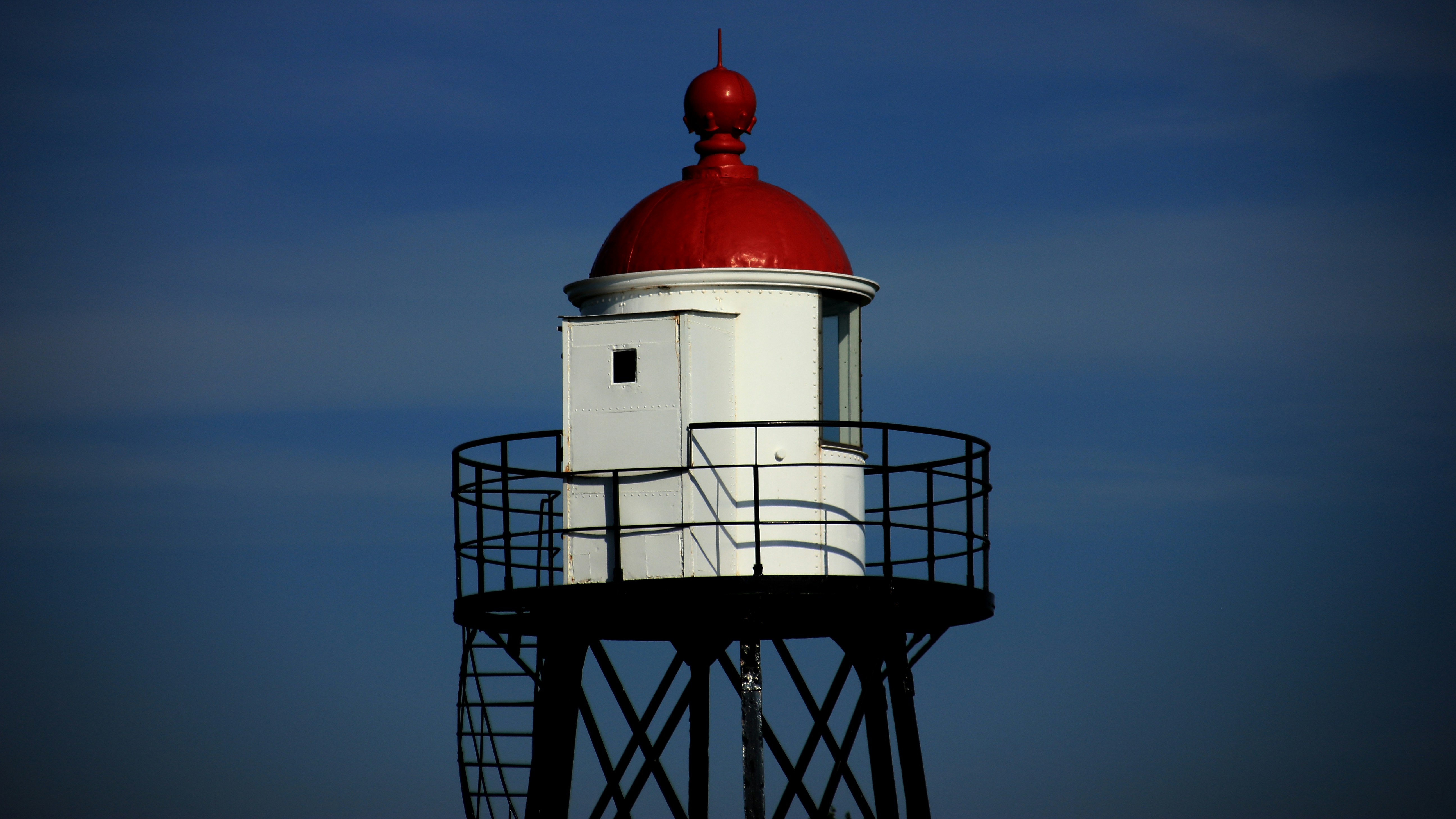 White lighthouse with a red dome atop a metal framework against a deep blue sky.