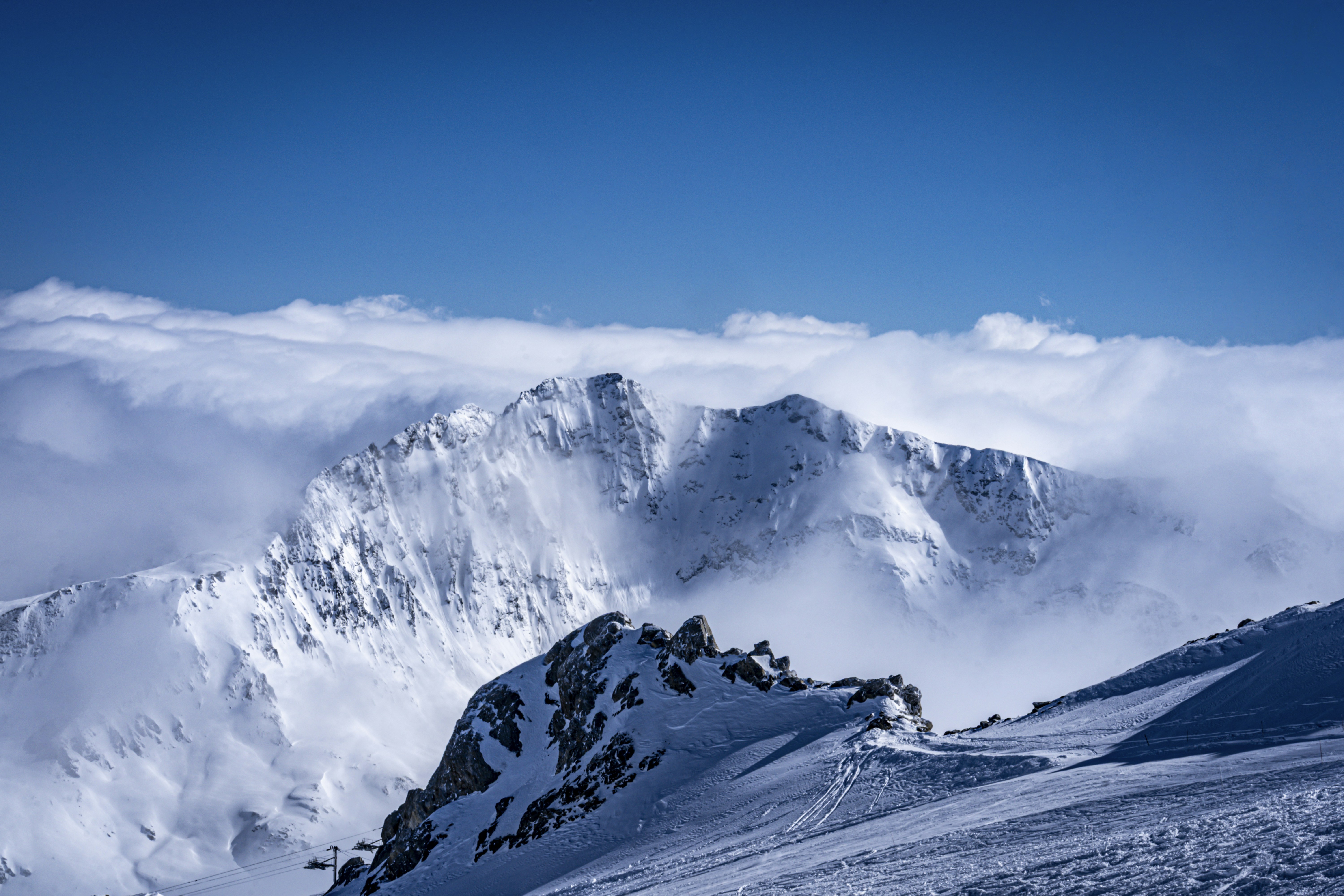 Snow-capped mountains emerge through dense clouds under a vivid blue sky.