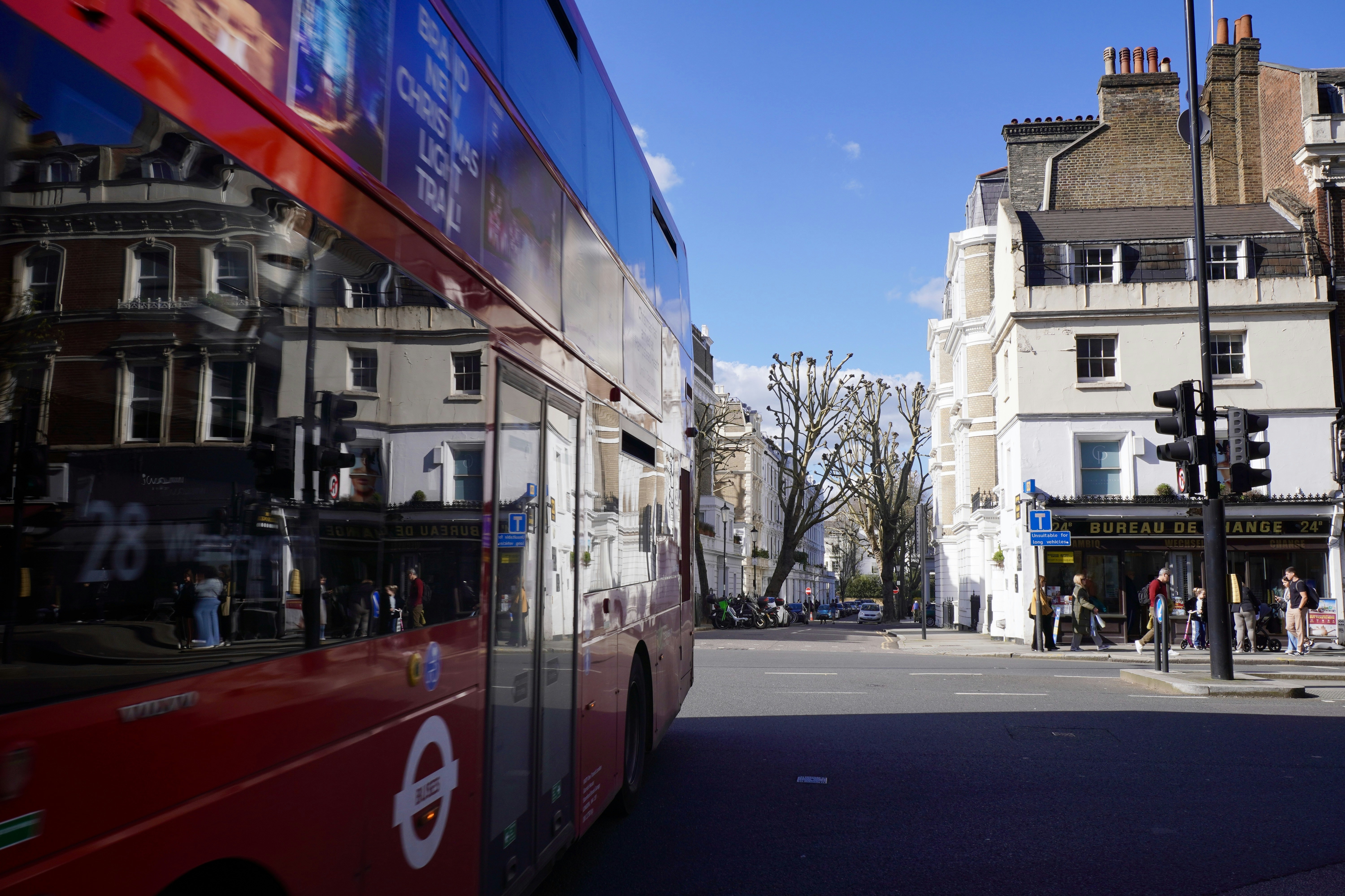 Red double-decker bus at an intersection near Notting Hill Gate, reflecting historic buildings. Bright blue sky and white townhouses contrast the scene.