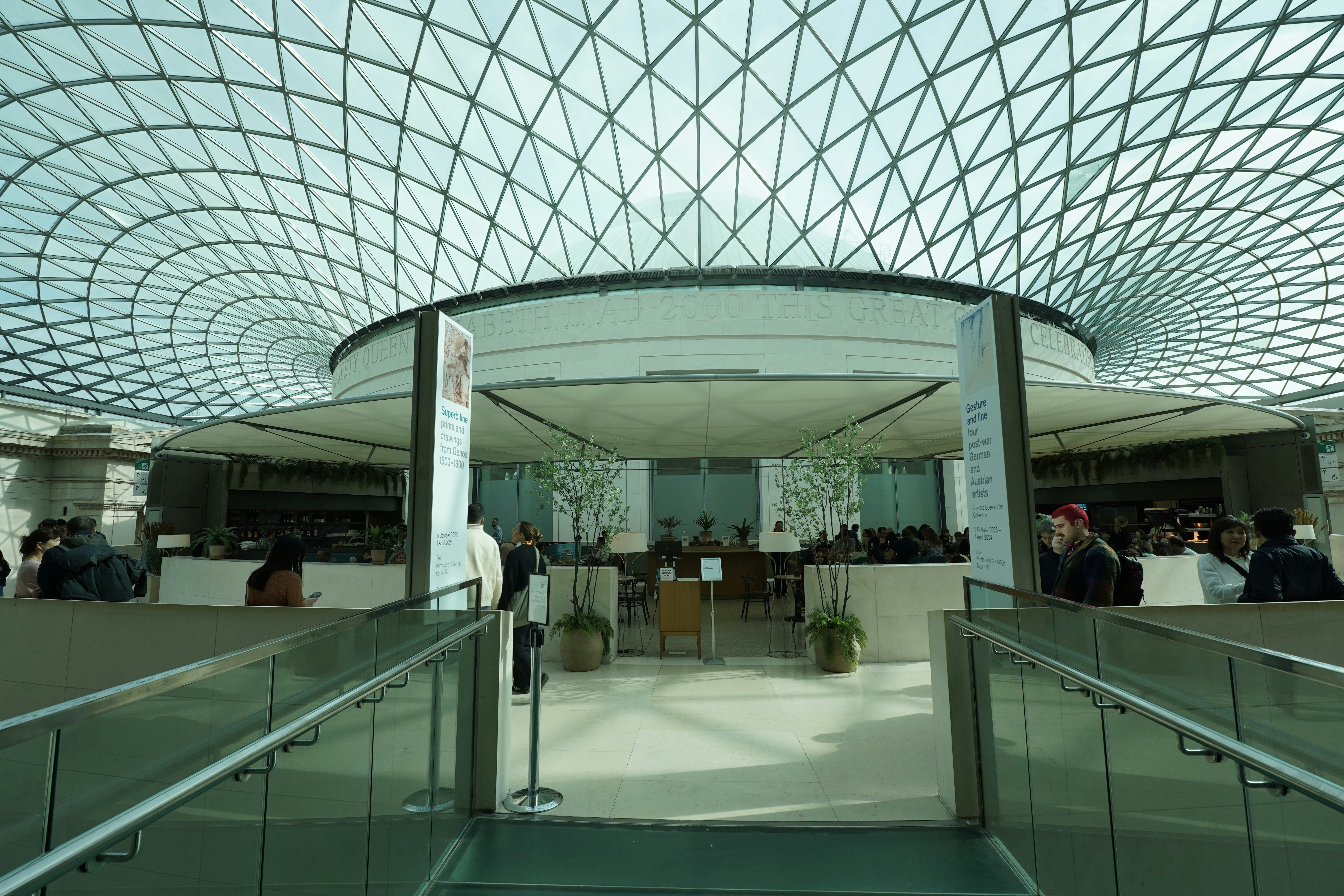 The Great Court Restaurant at the British Museum sits beneath the breathtaking glass and steel roof designed by architect Norman Foster. The geometric patterns of the ceiling create a dynamic interplay of light and shadow, while visitors enjoy refreshments in this iconic cultural space. A blend of history and modern design, the restaurant provides a unique atmosphere in the heart of one of the world's most famous museums. | A group of people are standing in a lobby