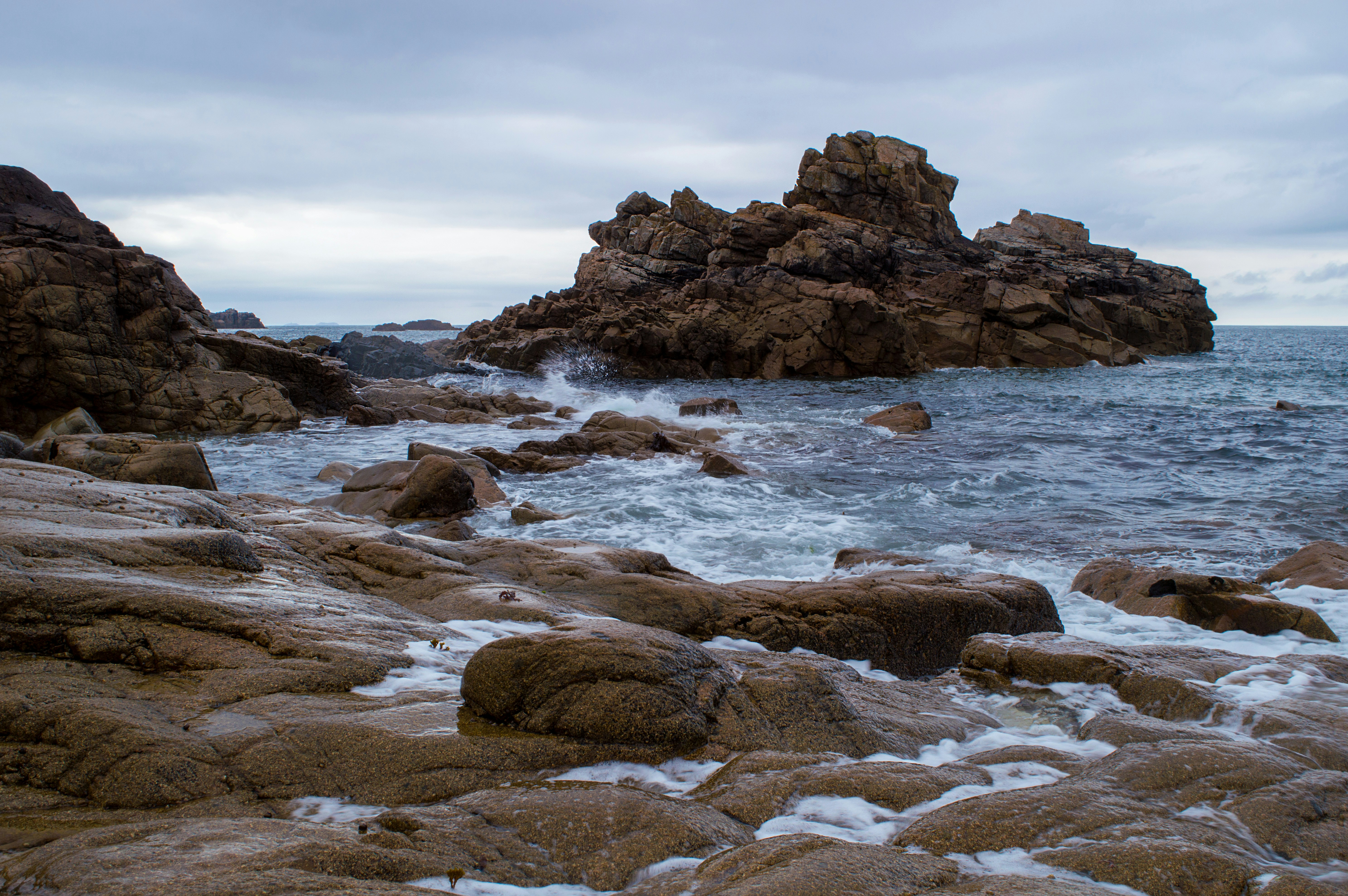 Massive brown rocks jut from the turbulent sea as waves crash, creating white foam under an overcast sky.