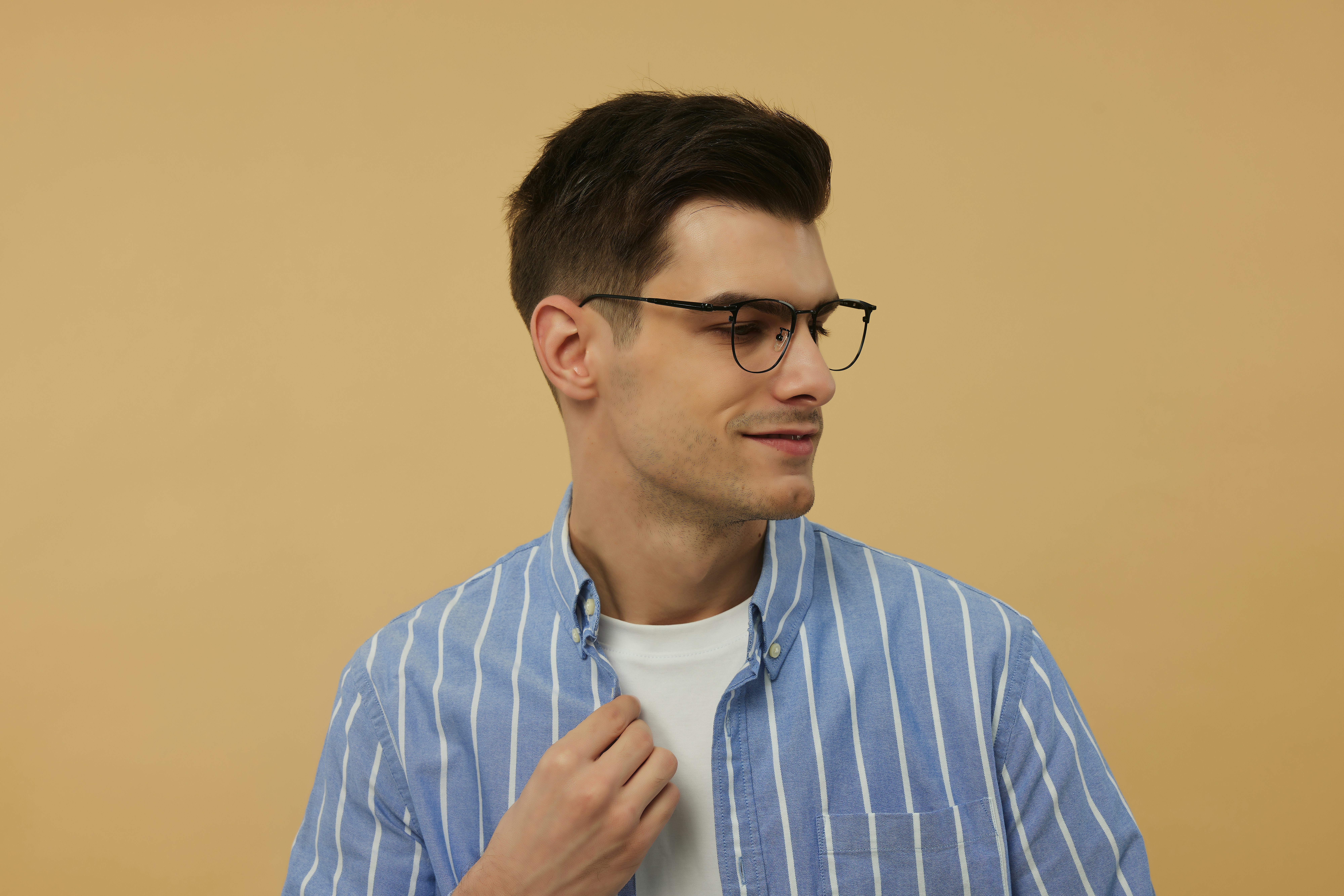 Young man with glasses and a striped shirt, looking thoughtfully to the side against a warm beige background.