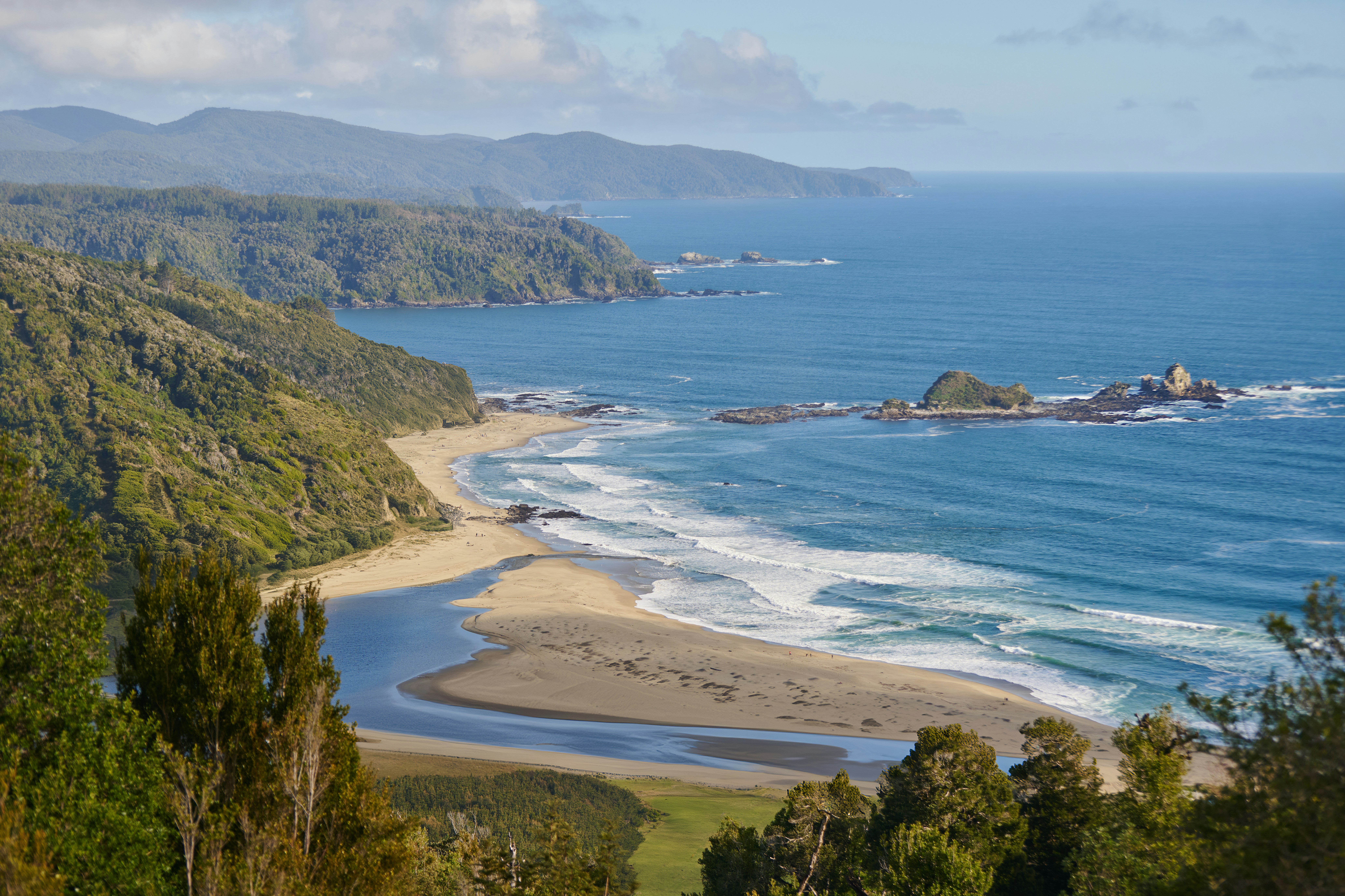 Coastal landscape with winding beach and lush green hills meeting the ocean under a partly cloudy sky.