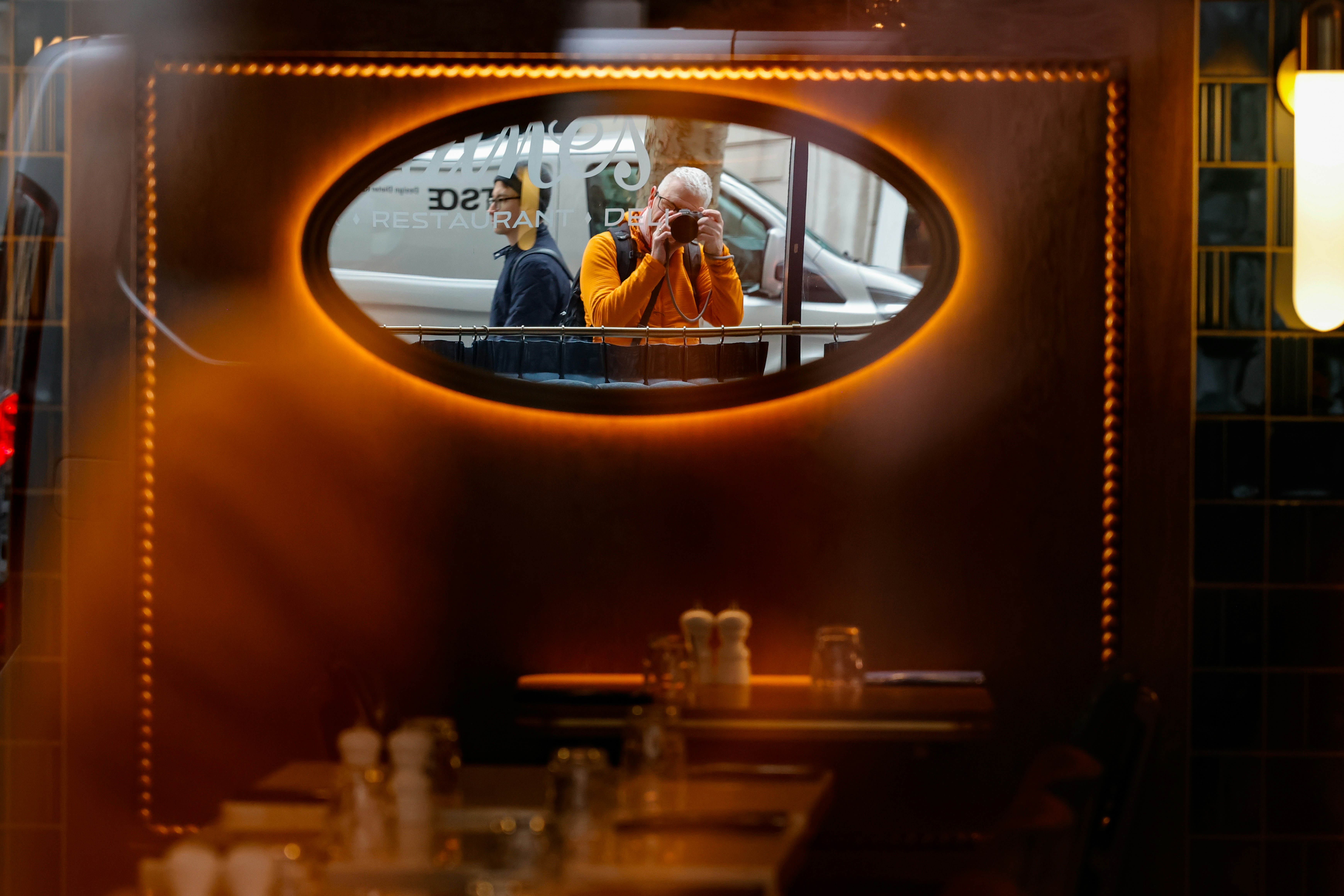 Photographer's reflection captured in an oval window of a warmly lit café interior.