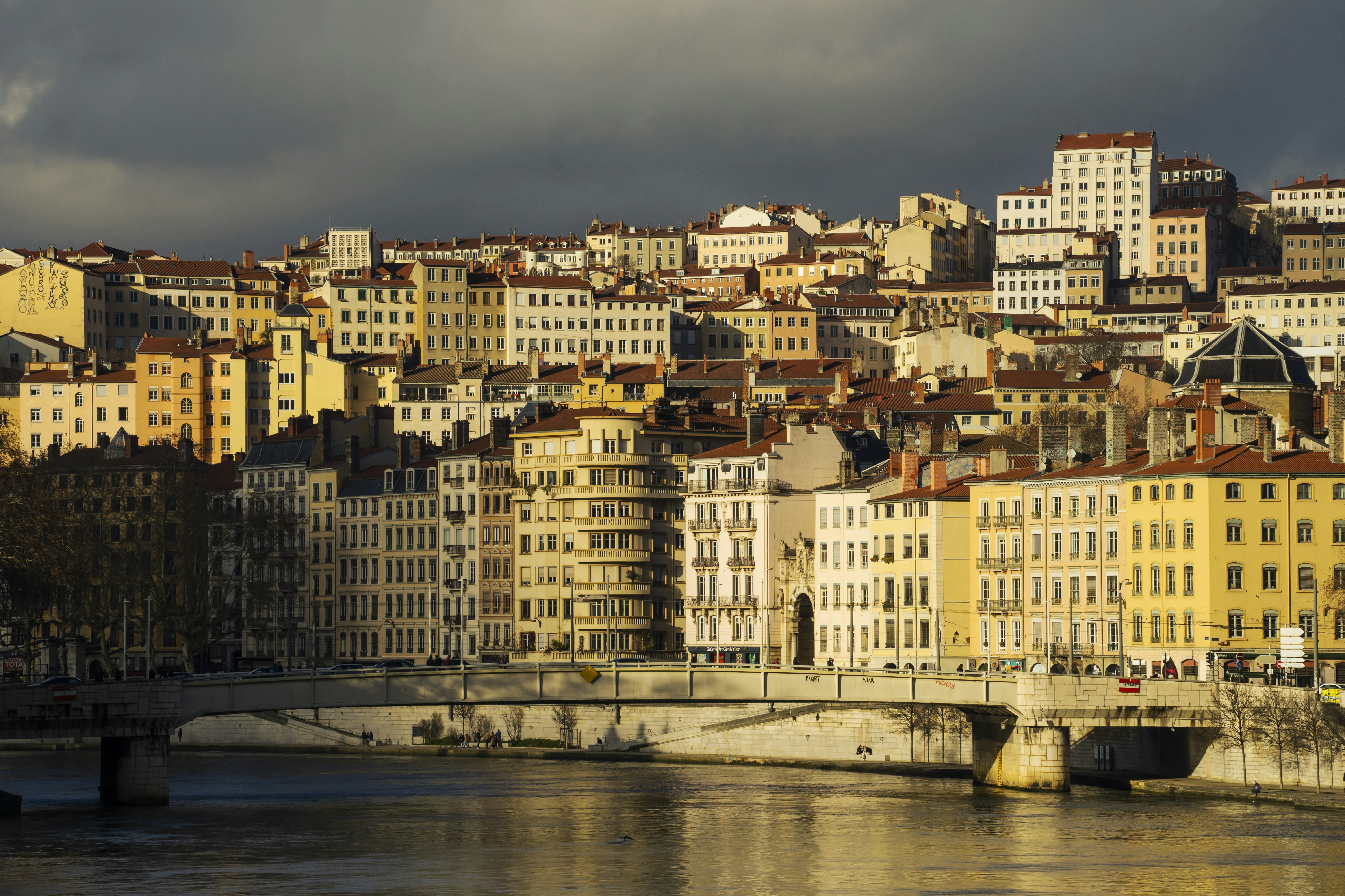 Sunlit buildings by the river, showcasing varied architectural styles under a dramatic sky.