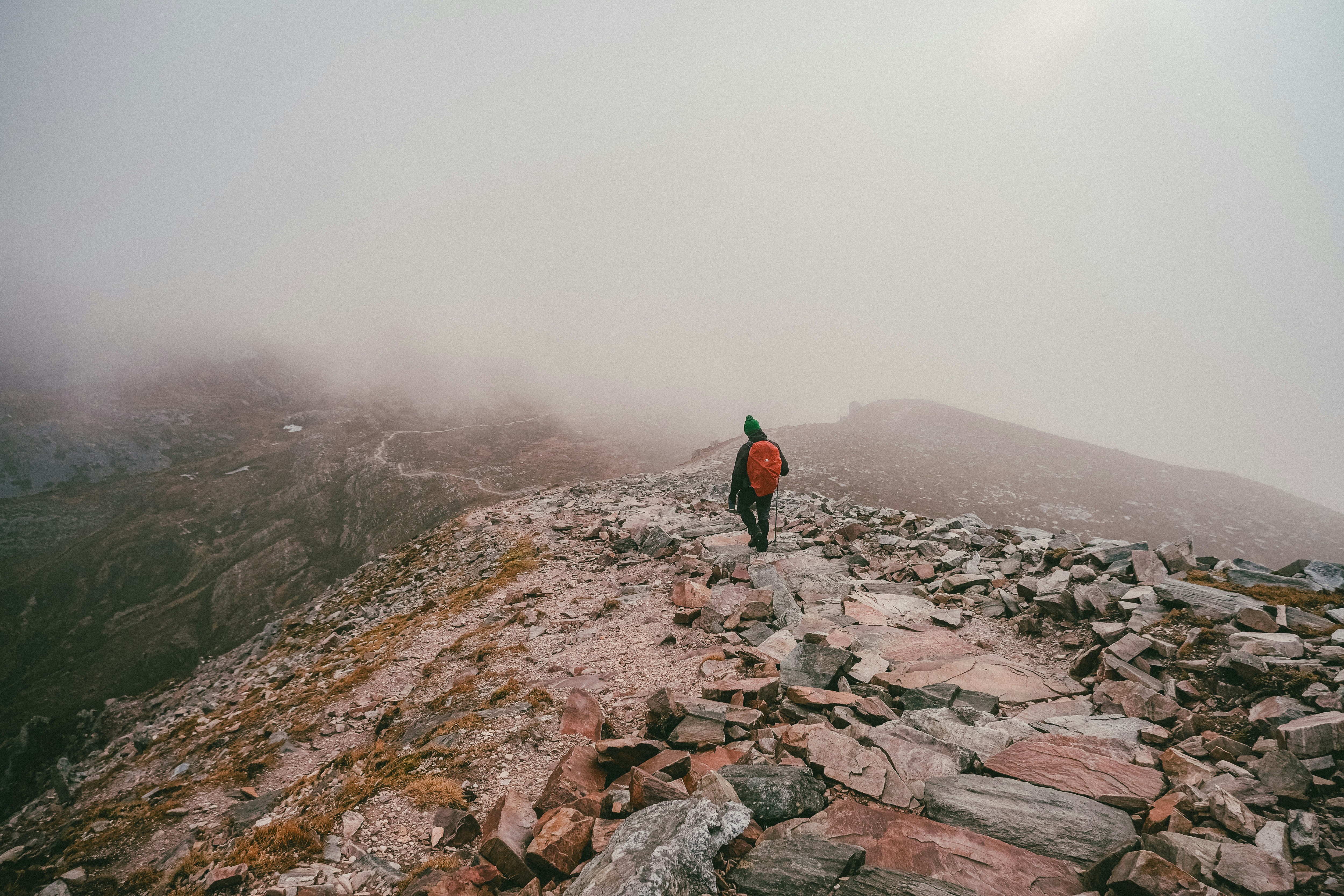 Hiker with a red backpack walking along a rocky mountain ridge shrouded in fog.