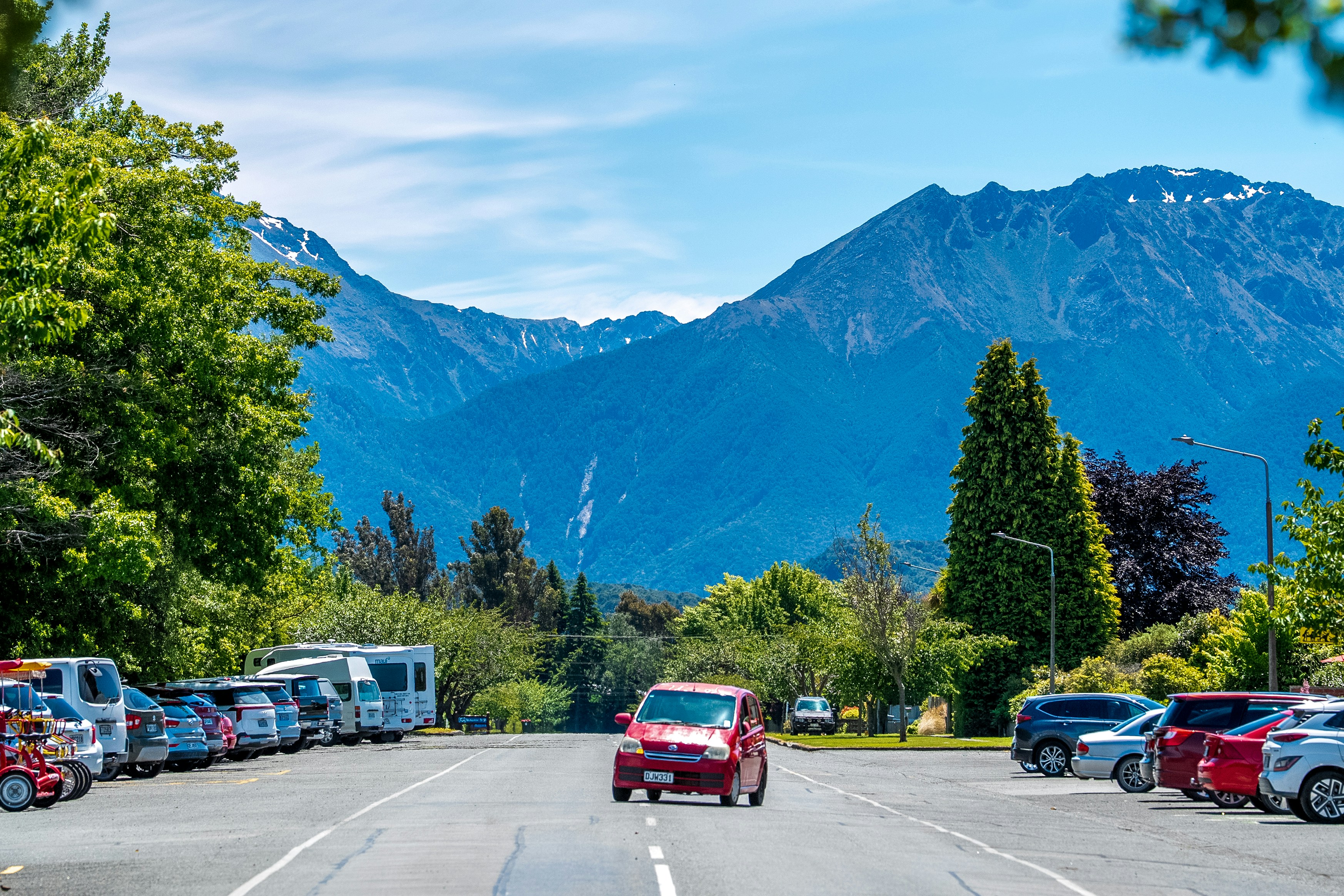 Red car travels down a tree-lined road with towering mountains in the background under a clear blue sky.