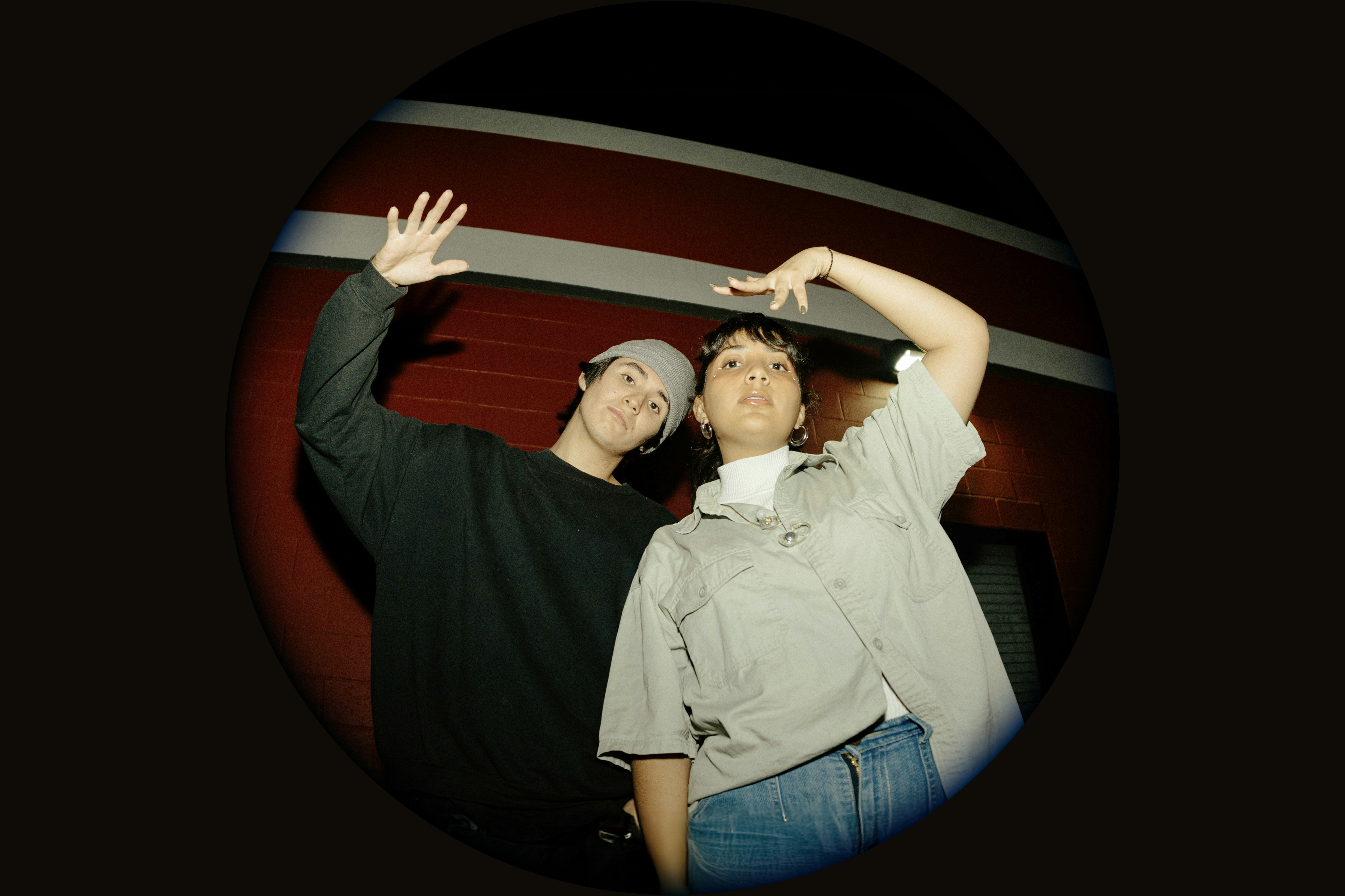 Two individuals pose expressively against a brick wall under artificial light.