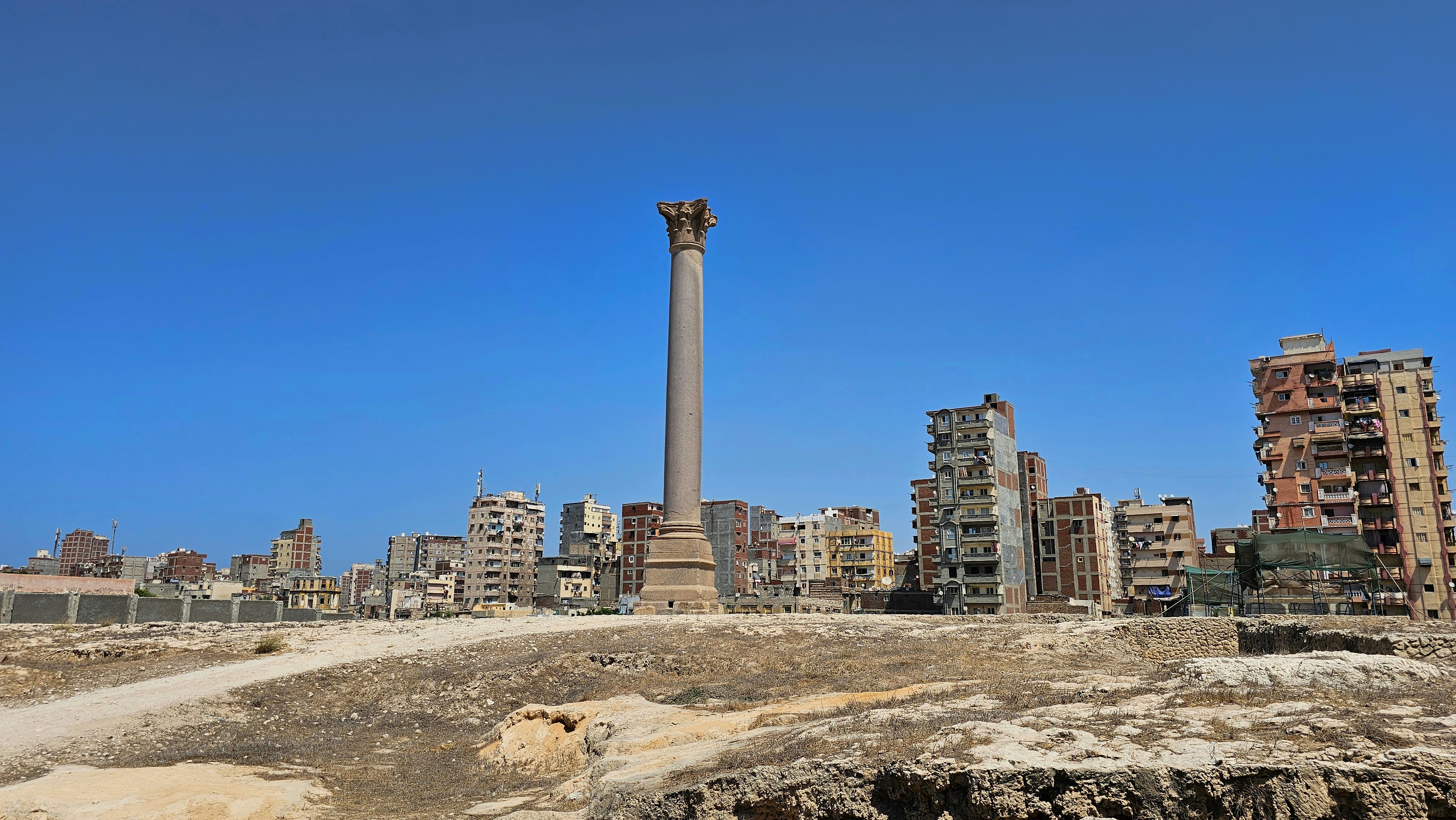 Ancient column standing amidst modern cityscape under a clear blue sky.