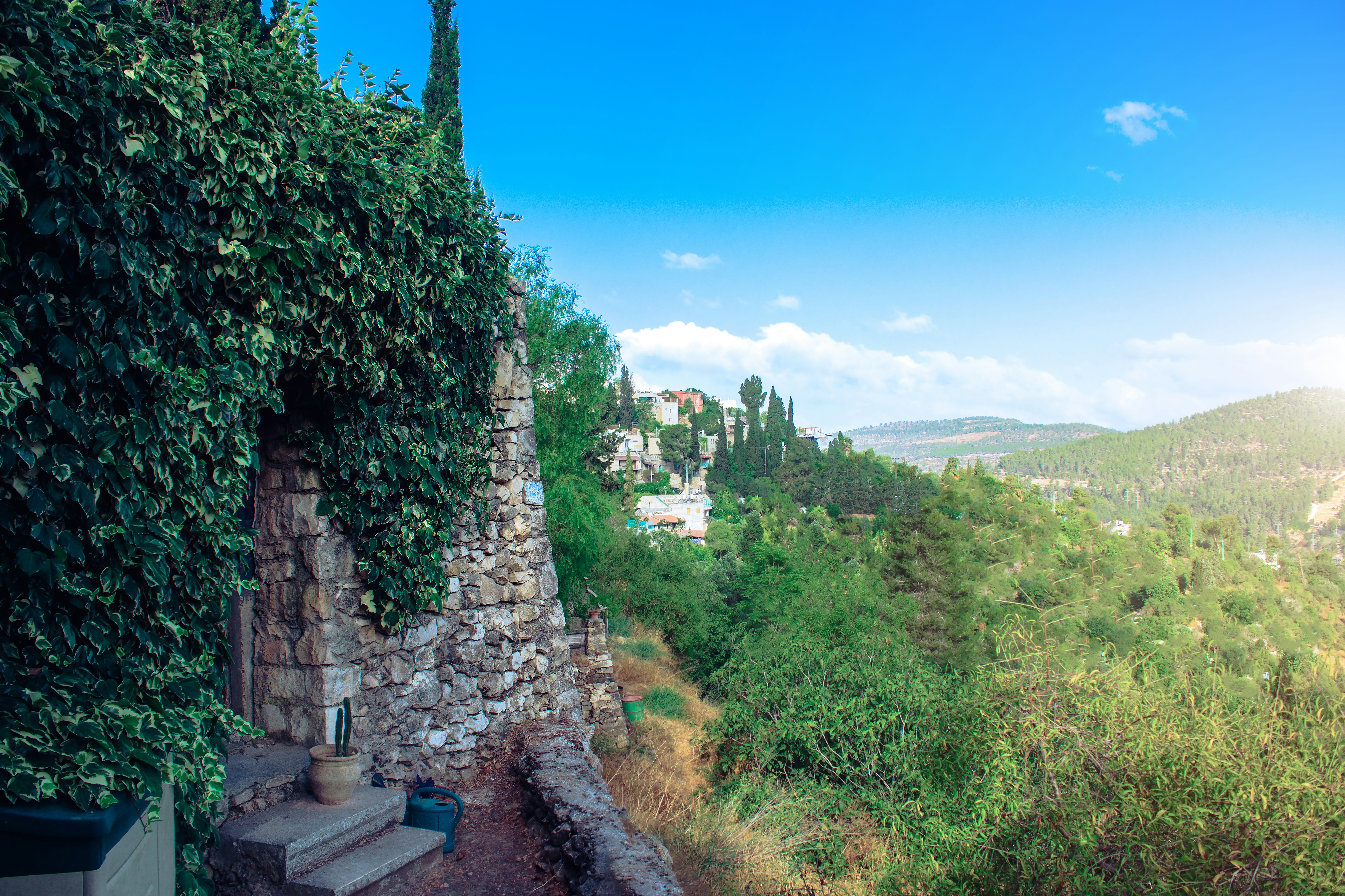 Stone farmhouse entrance draped in lush greenery with a view of rolling hills and distant village under a clear blue sky.
