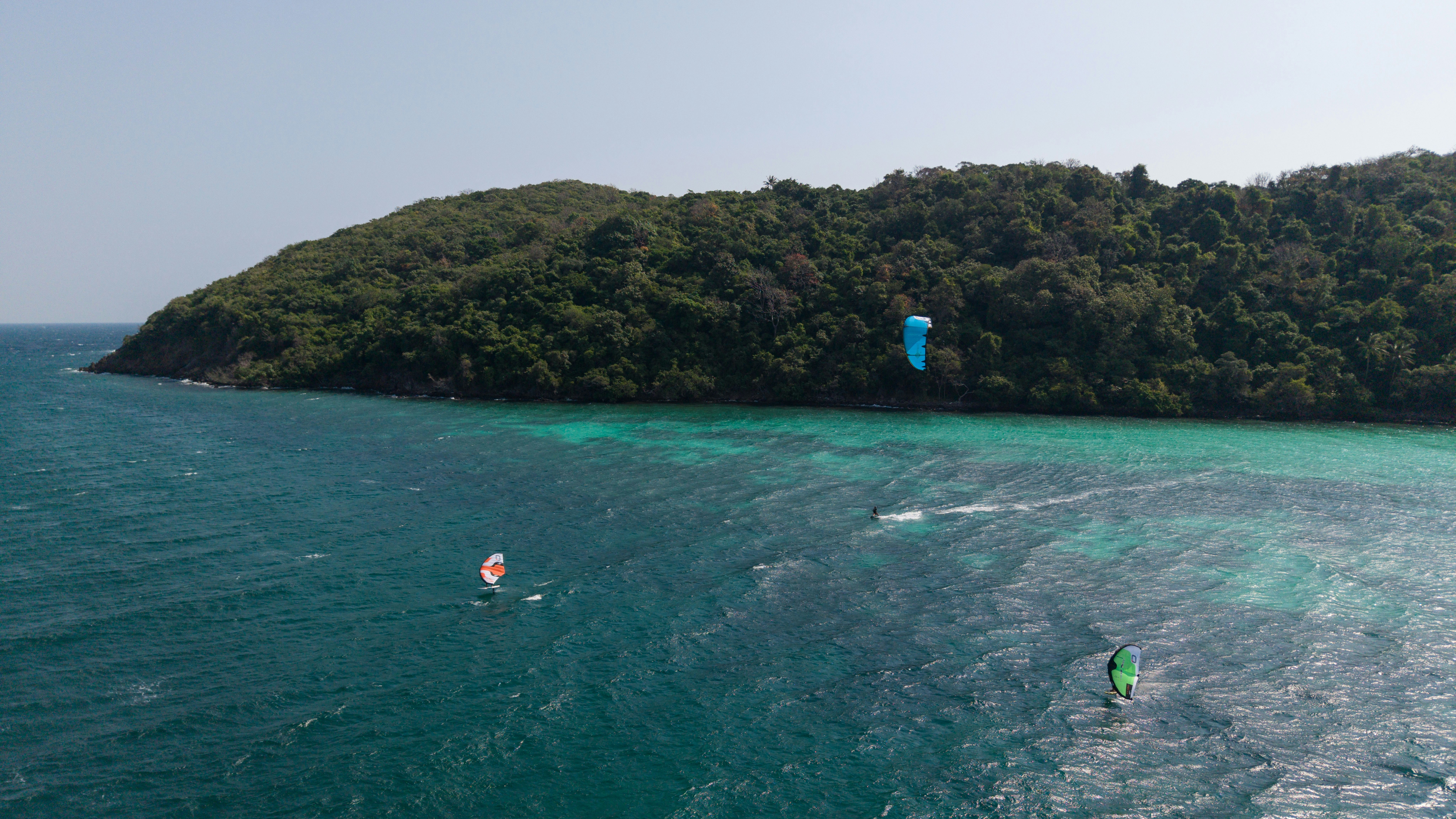 Kitesurfers glide across vibrant turquoise waters near a lush, forested island.