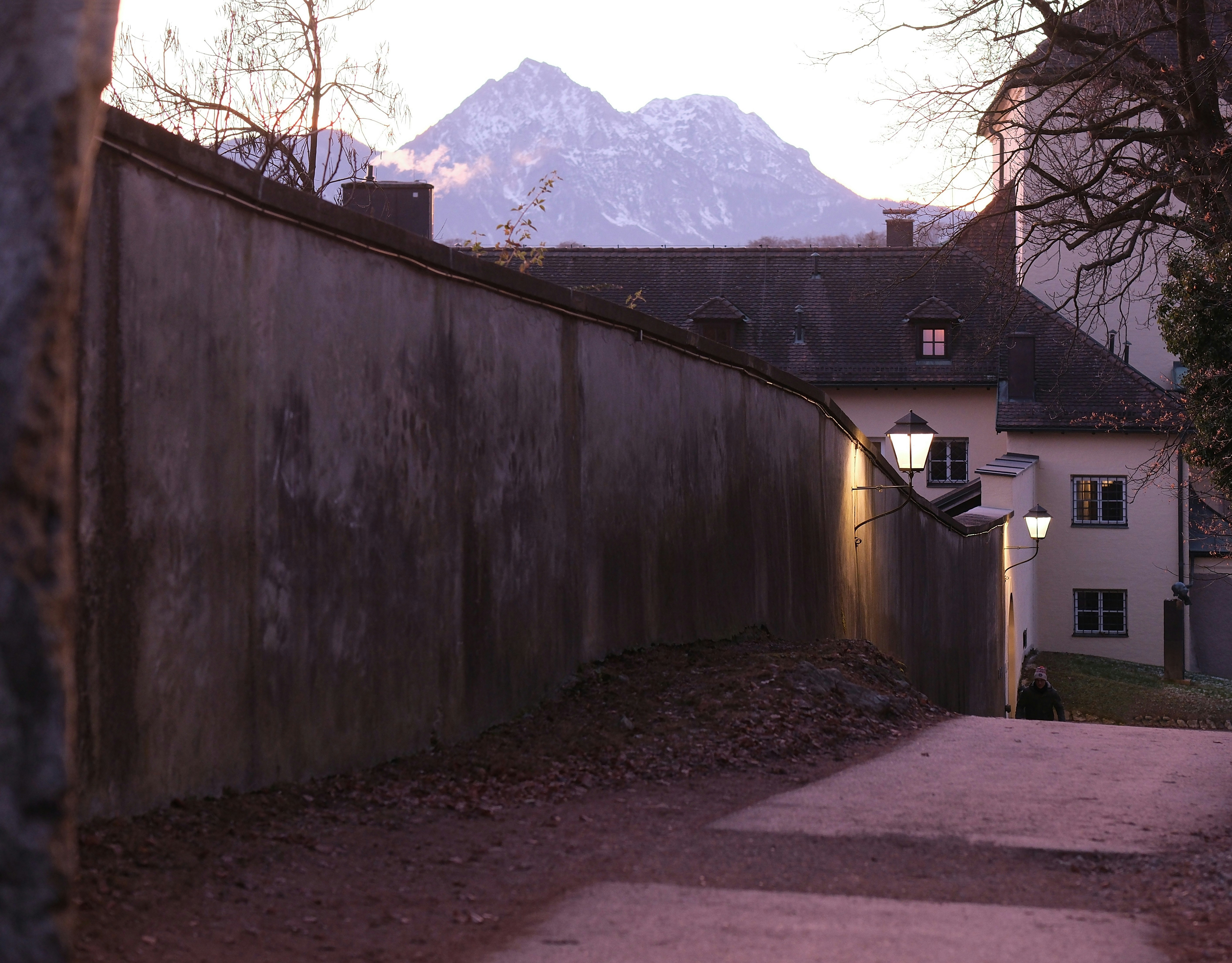 Dusk light casts a serene glow over a narrow path flanked by old Salzburg houses with distant mountains.