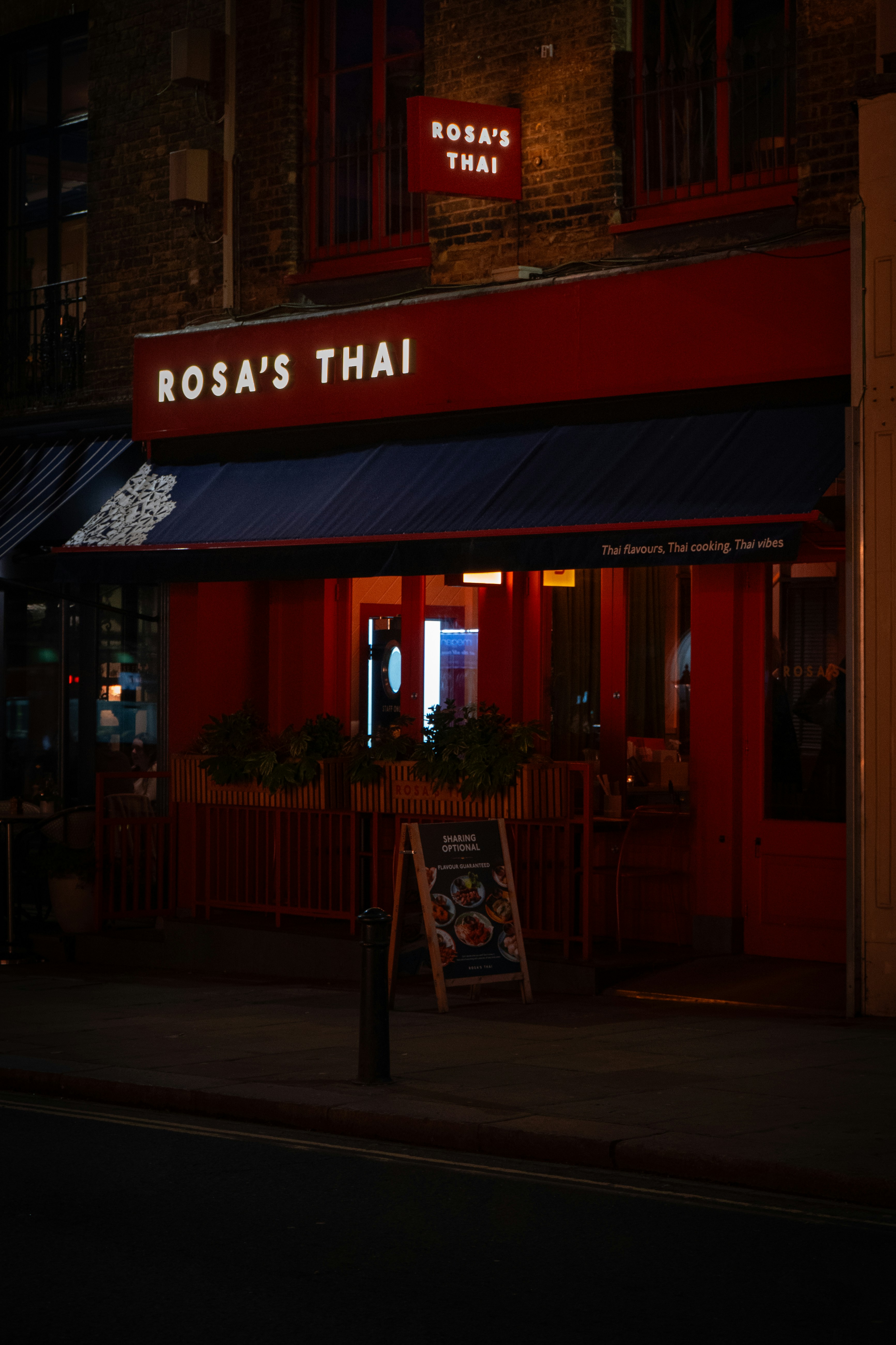 Streetfront of Rosa's Thai with warm red and blue tones under dim evening light.
