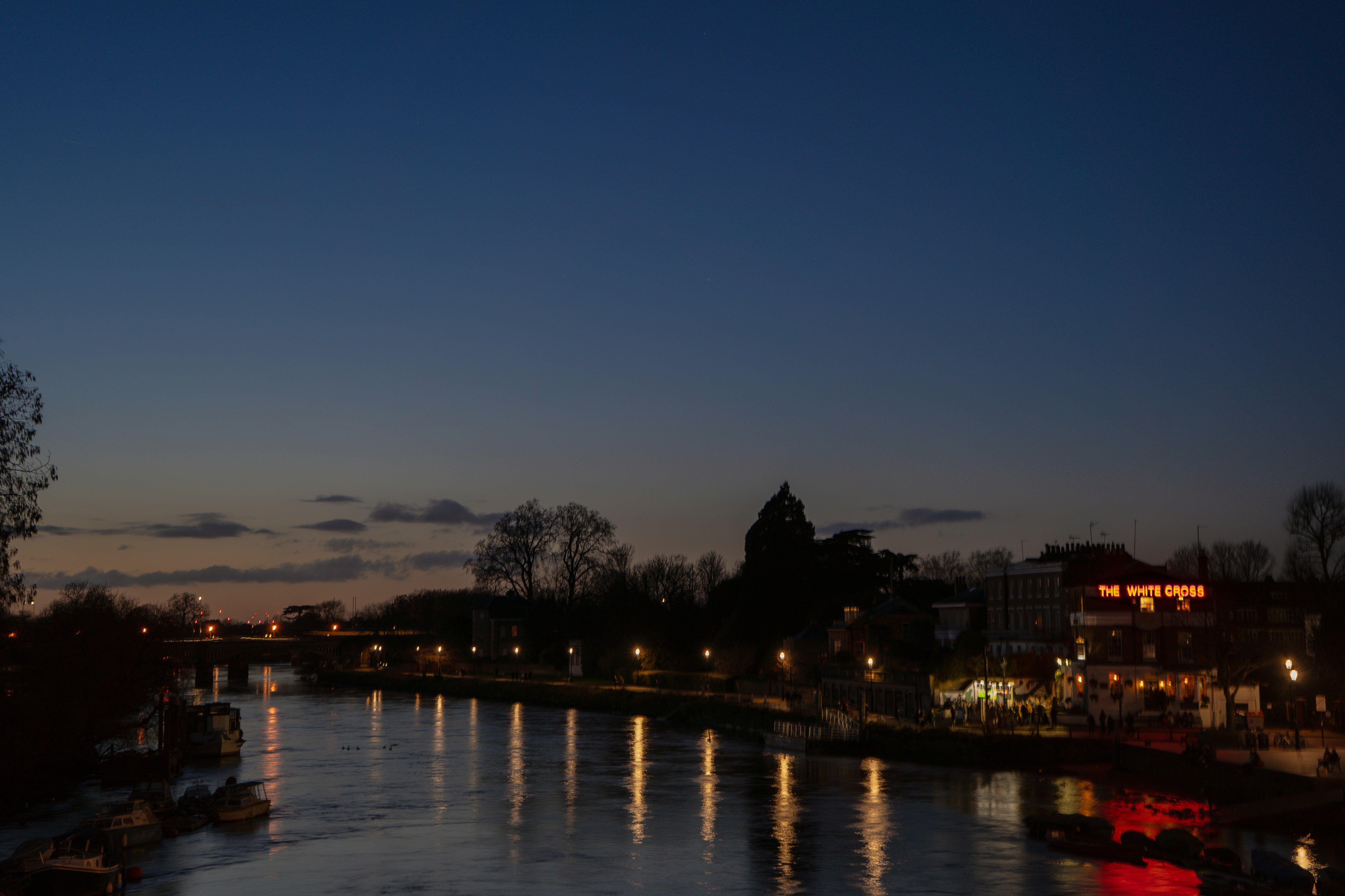 Dusk settles over a river with soft lights reflecting on the water, framed by trees and a distant skyline.