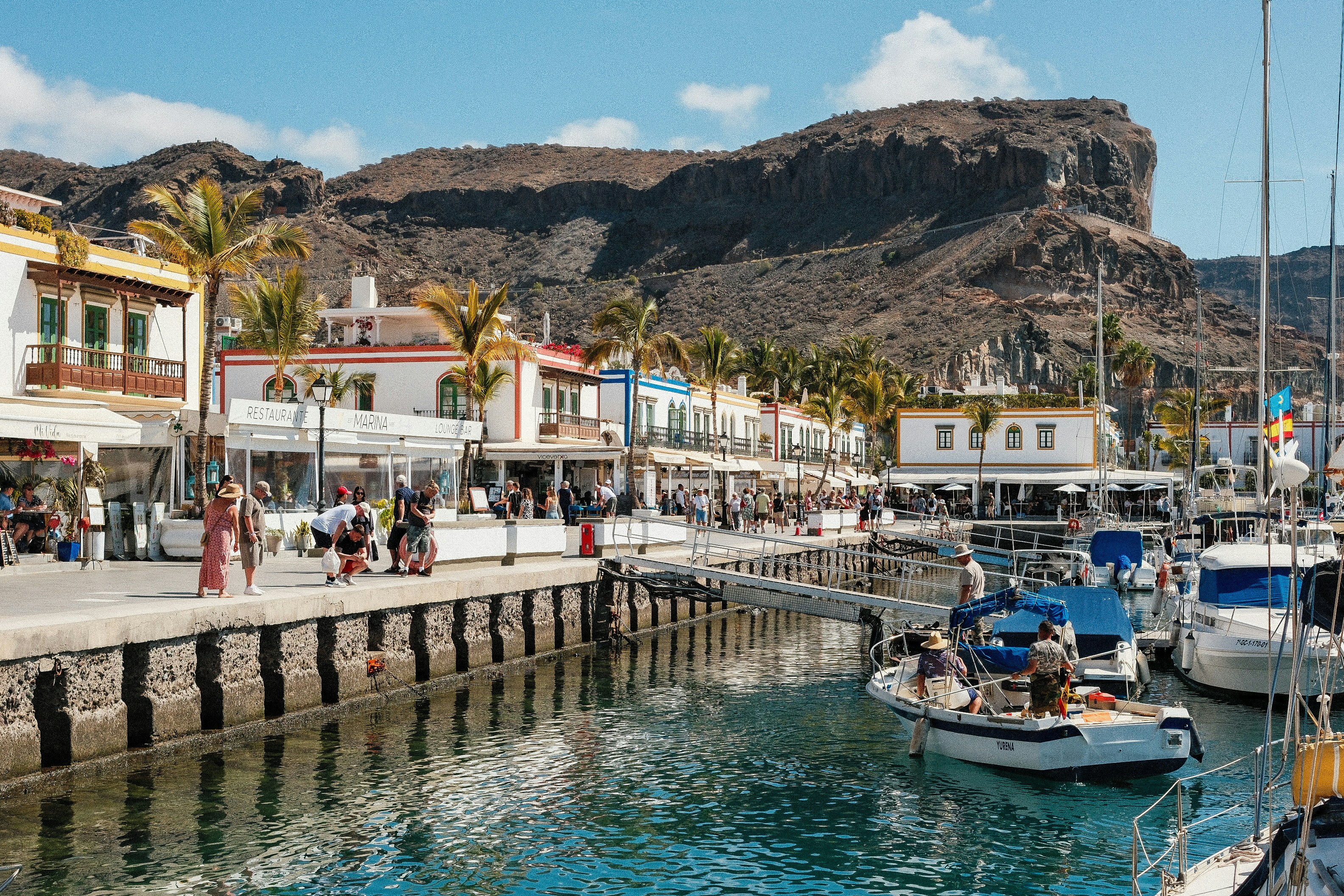 Vibrant waterfront promenade with colorful buildings and moored boats under a rocky hillside.