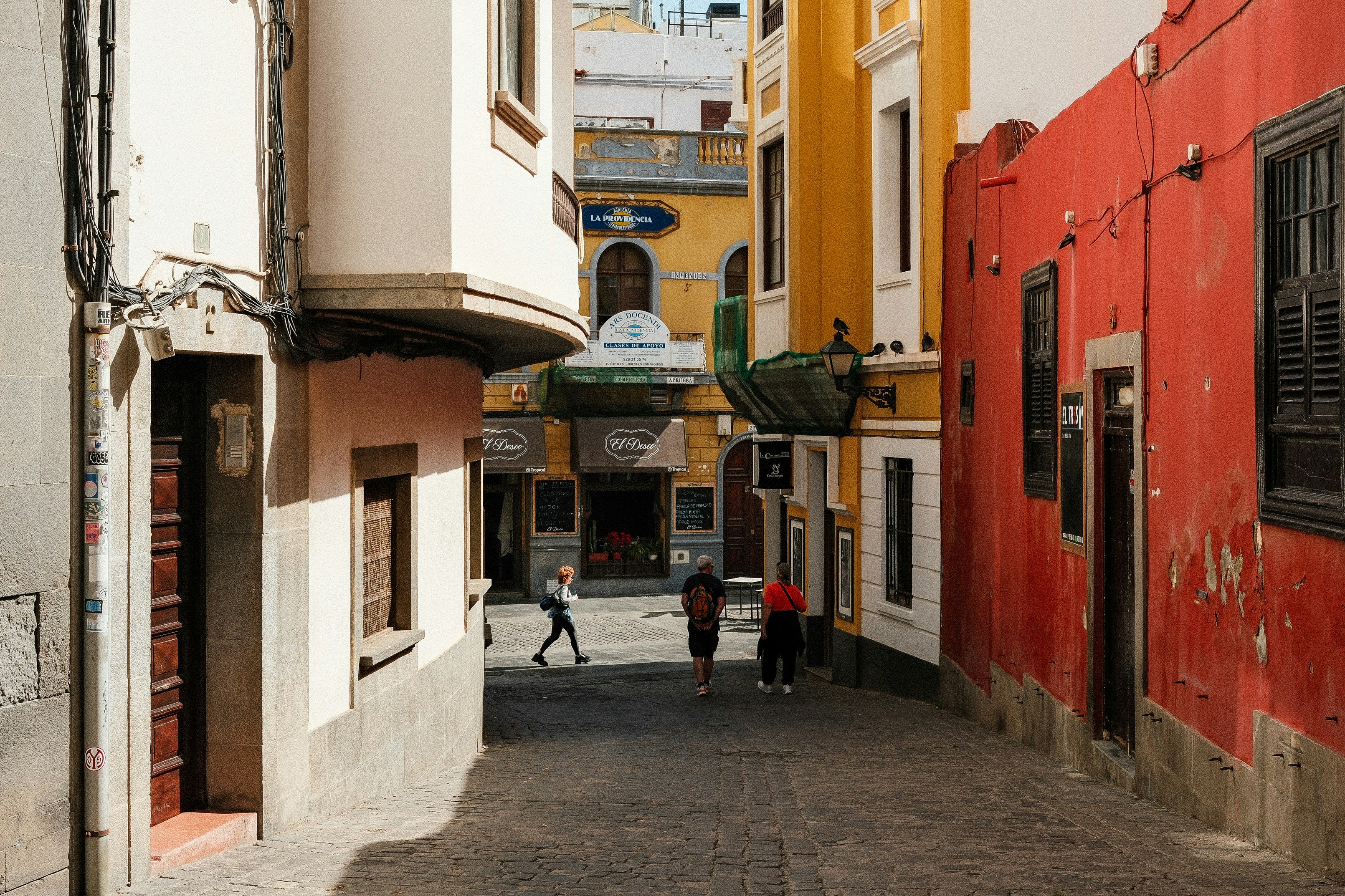 Narrow alley with vibrant yellow and red buildings, as pedestrians walk along sunlit cobblestones.