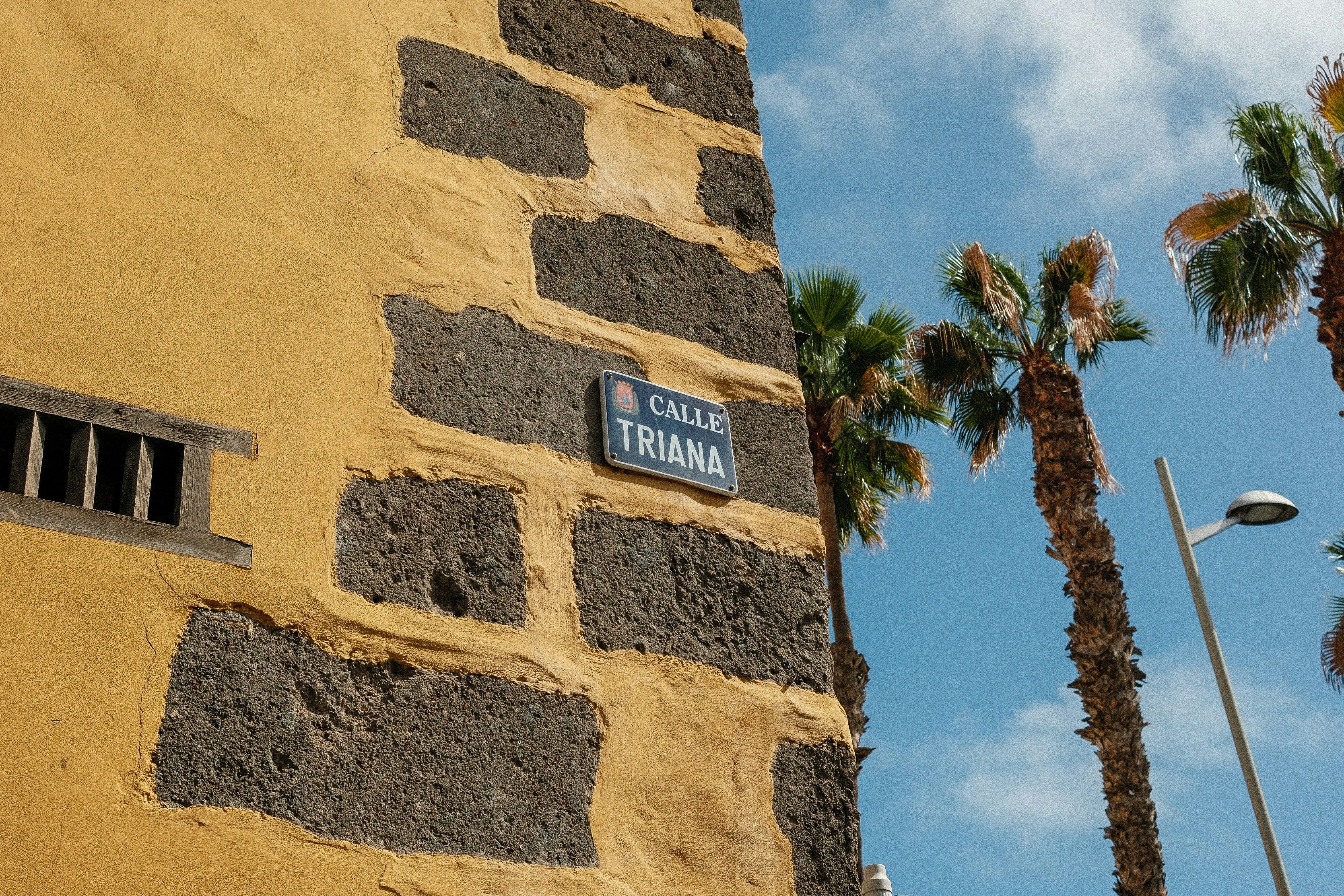 Street sign on a rustic stone building corner with palm trees swaying under a blue sky.