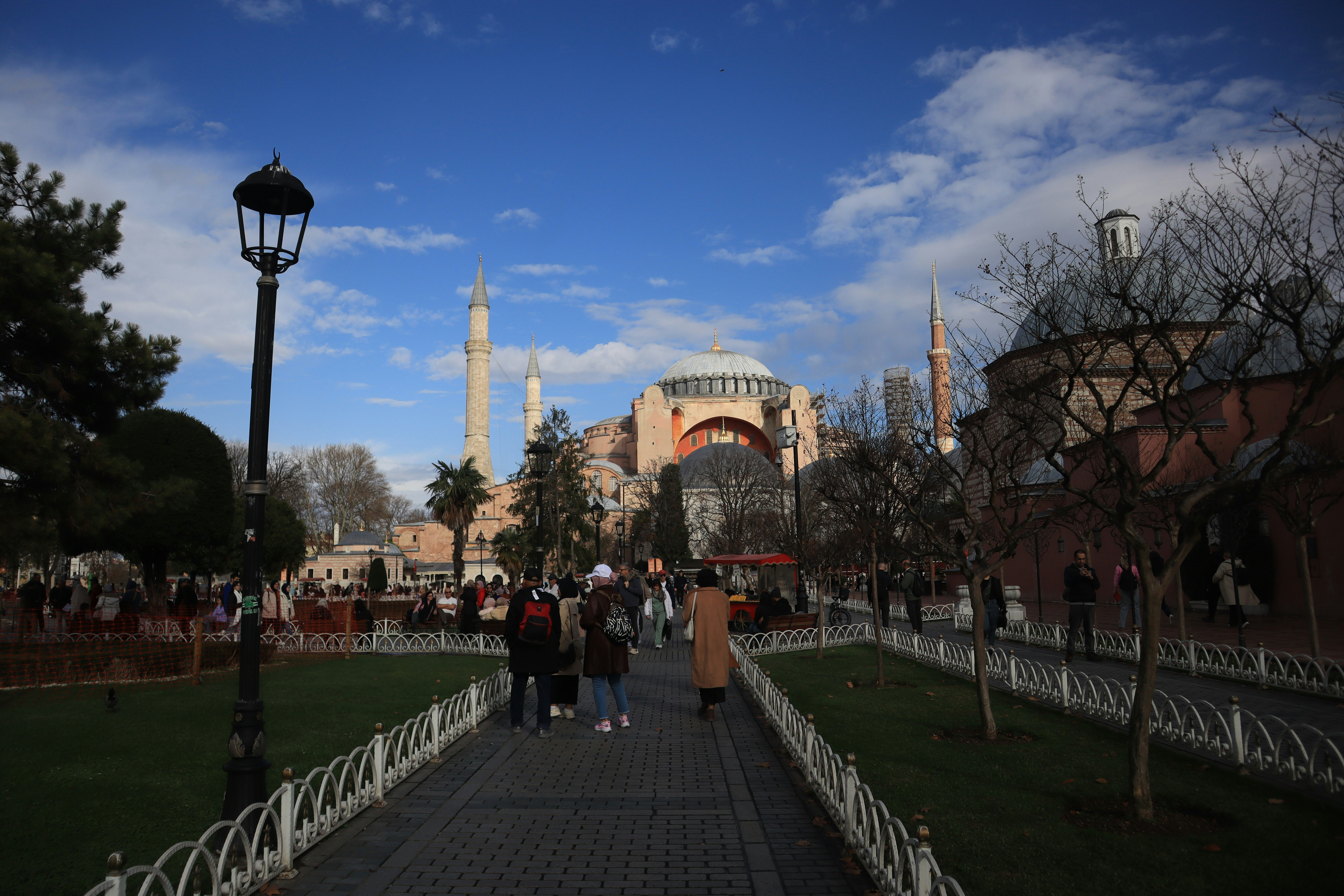 Hagia Sophia Grand Mosque under a vibrant blue sky, framed by a tree-lined walkway and distant minarets.