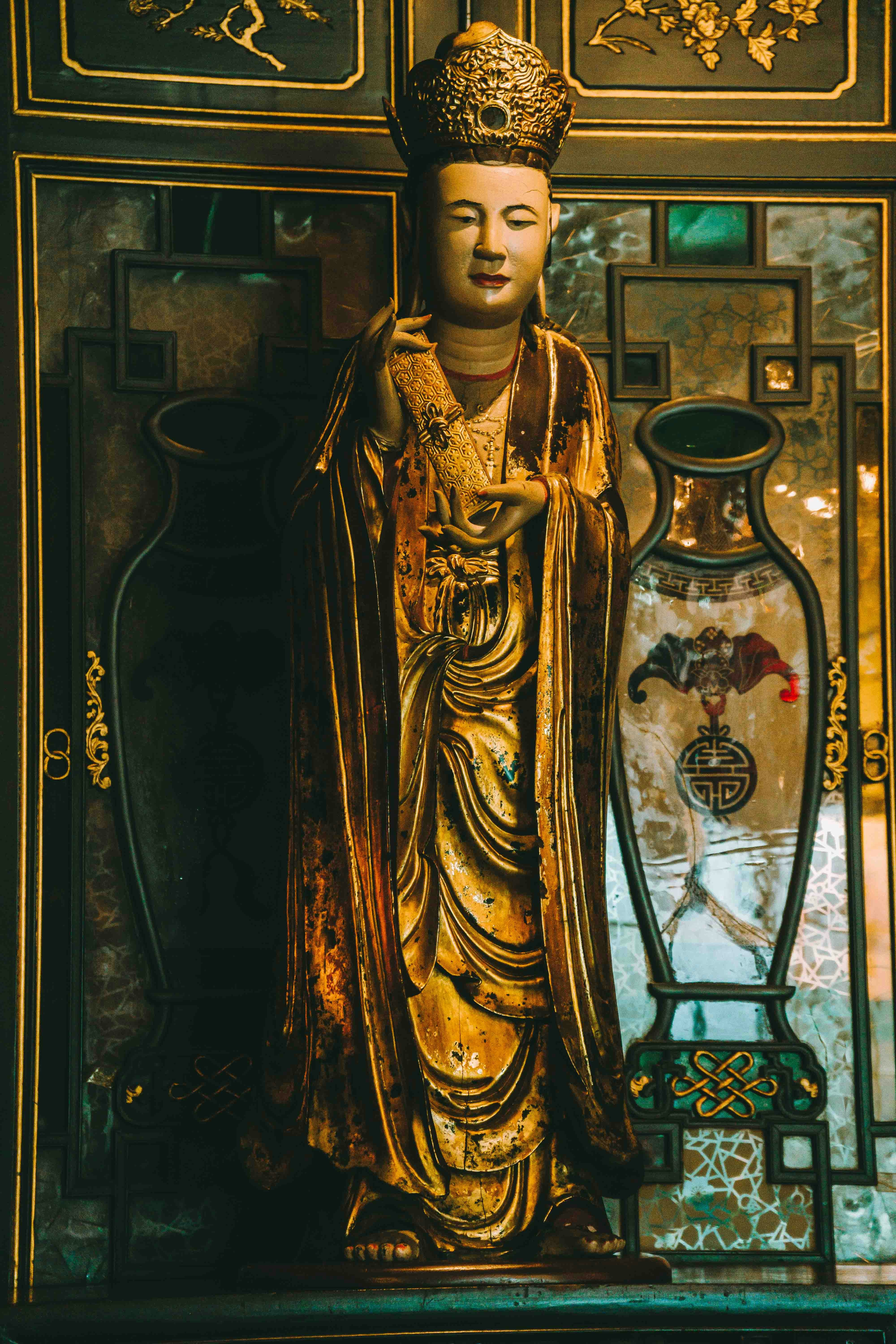 A gilded Guanyin statue bathed in warm amber light, set within an ornate cabinet of ceramic panels and vases.