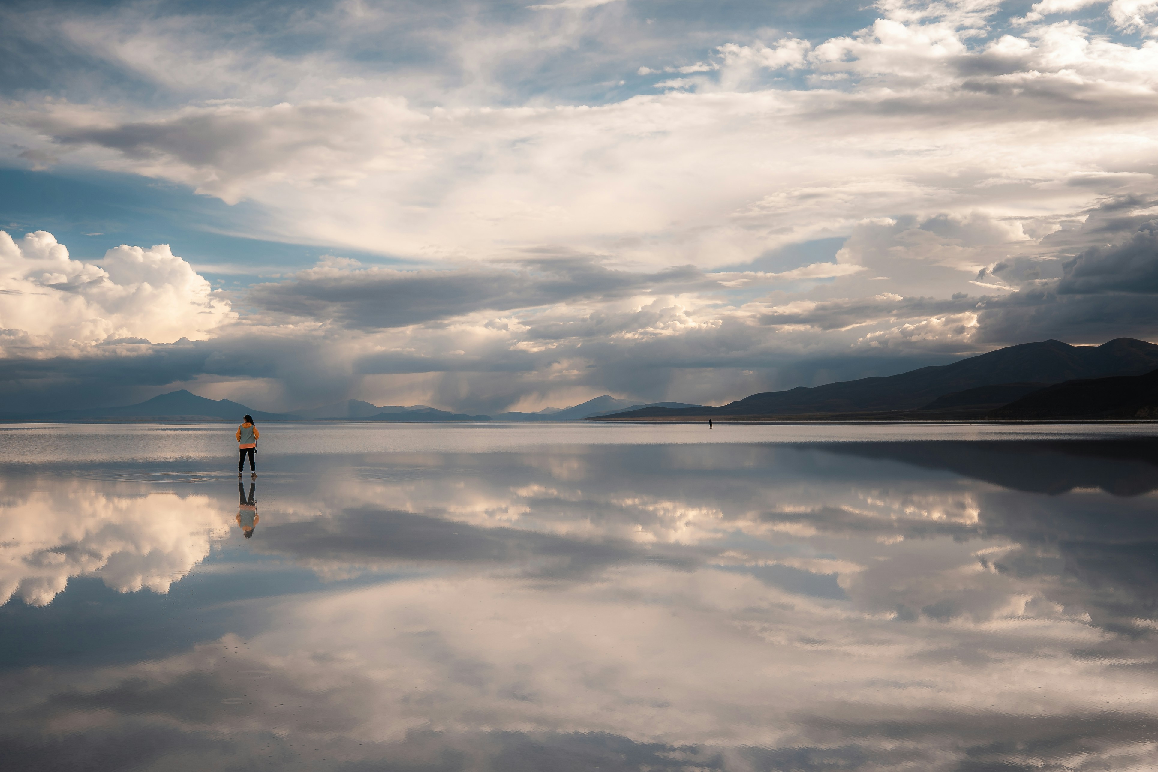 Person standing on vast reflective salt flat under dramatic cloudy sky.