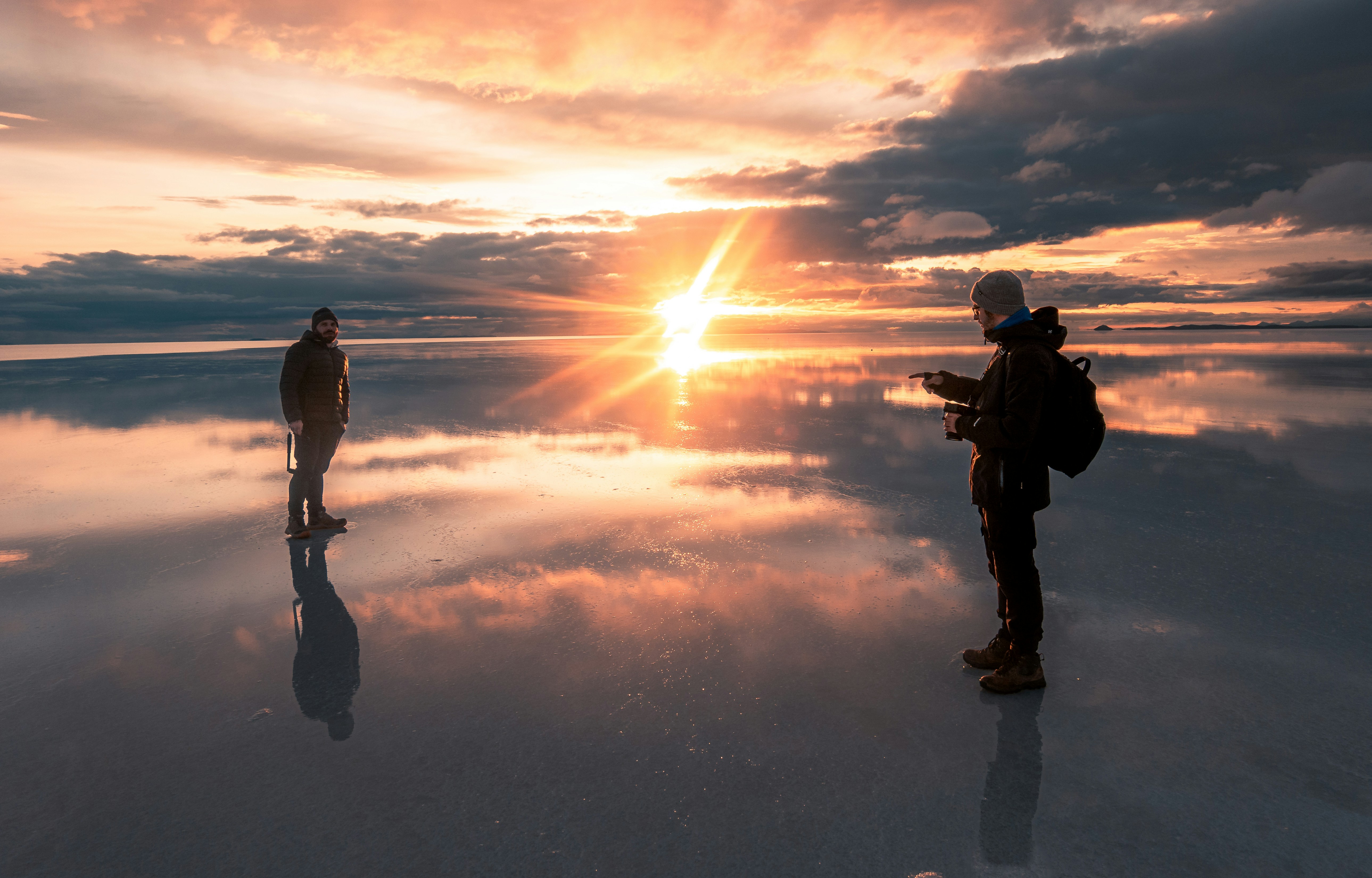 Two people standing on a reflective salt flat at sunset with vibrant clouds overhead.
