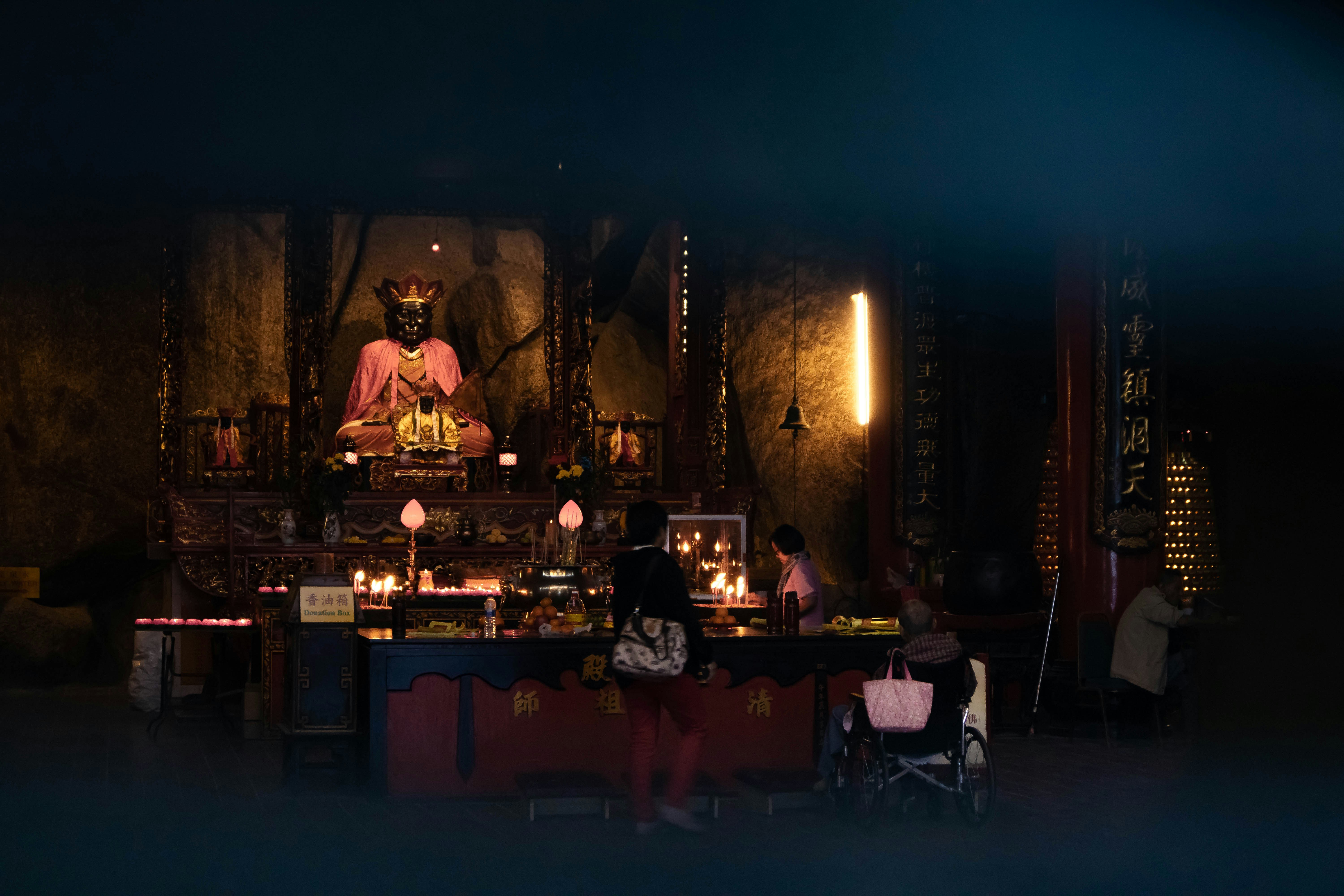 Dimly lit temple interior with a central Buddha statue surrounded by offerings and soft candlelight.
