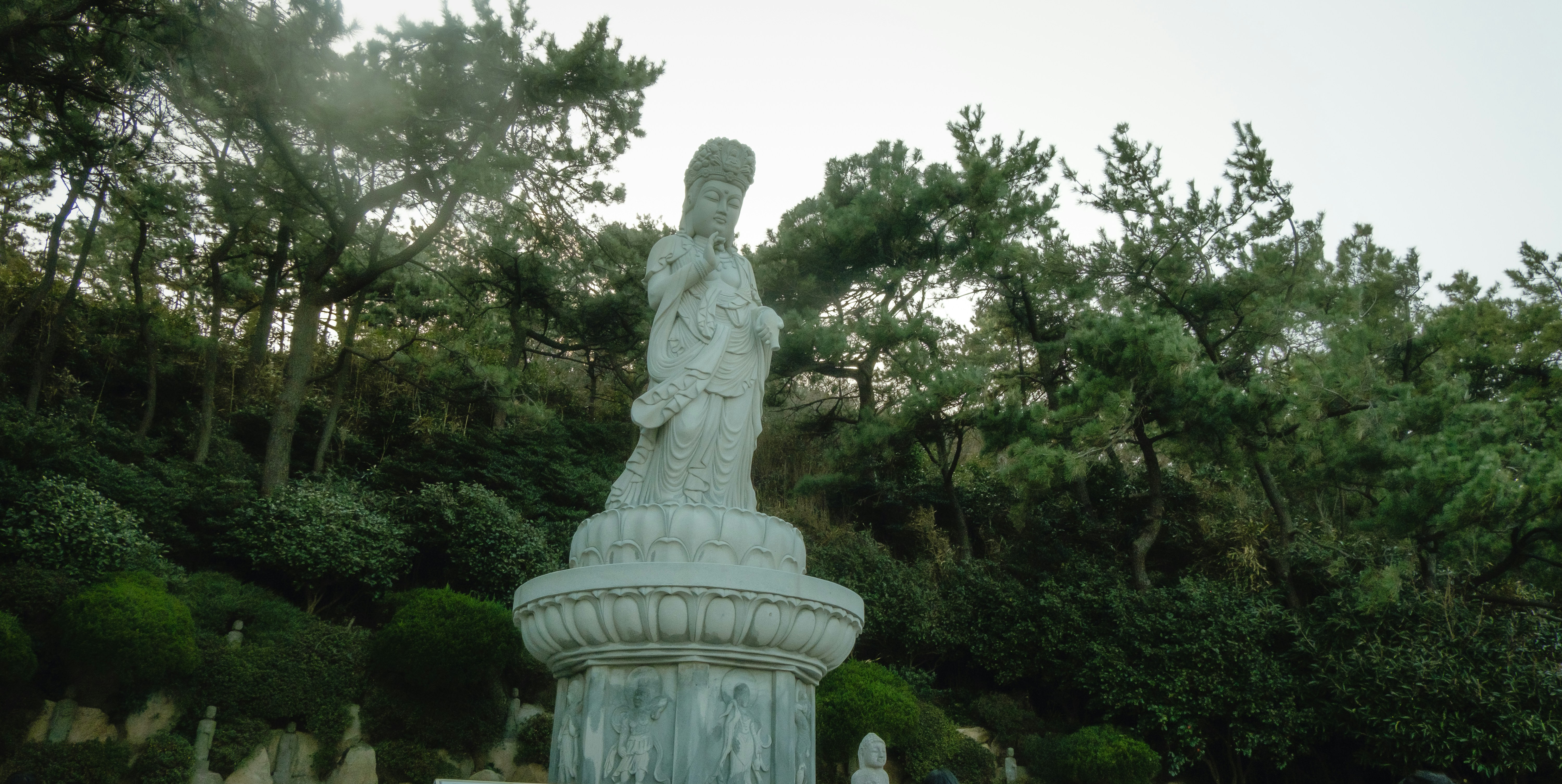Elegant stone statue nestled in a lush forest setting under a pale sky.