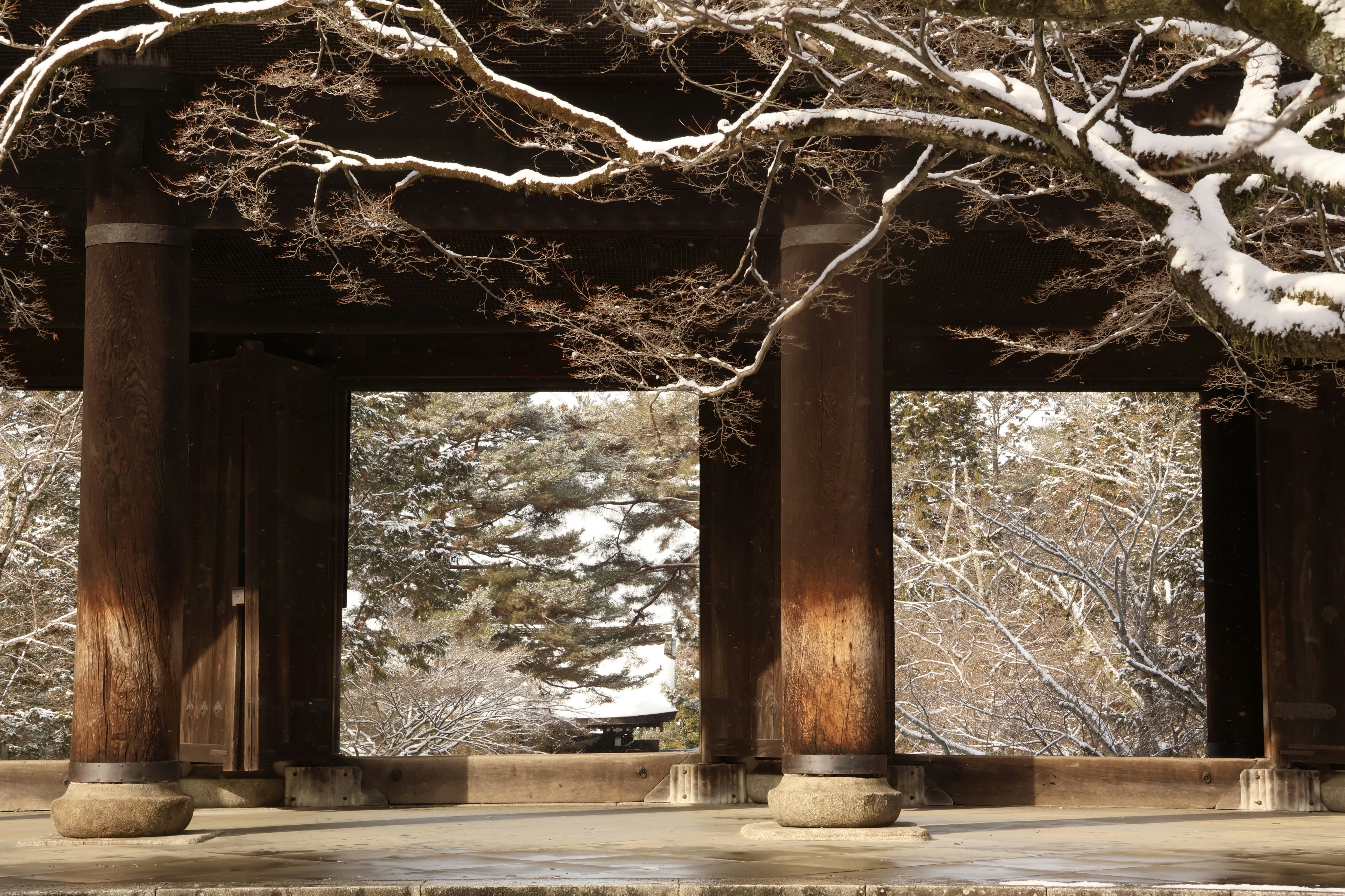 Snow-draped branches frame wooden pillars of a traditional structure, with a tranquil winter landscape beyond.