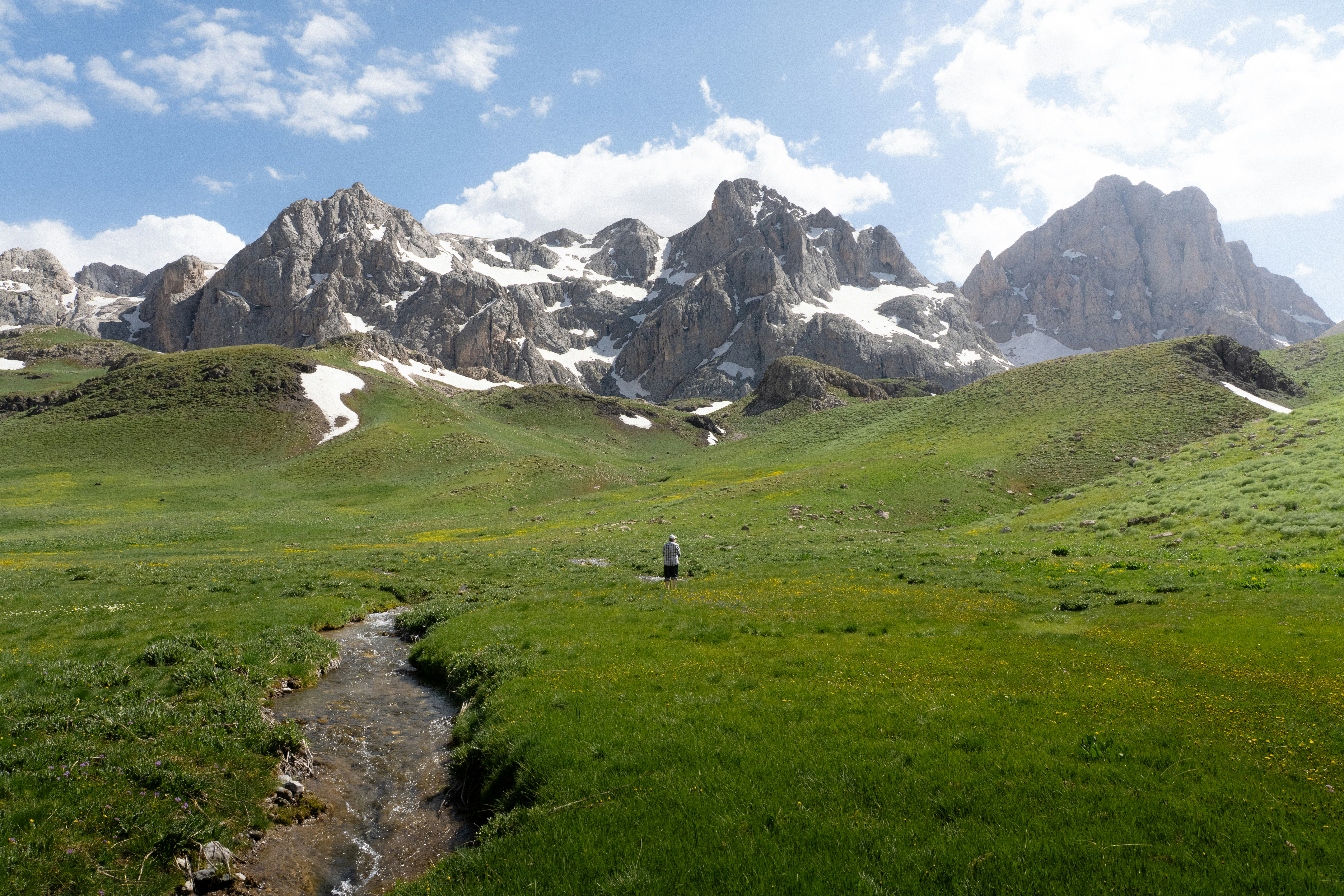 Mountains with snow patches loom over a lush green meadow and a meandering stream under a blue sky.