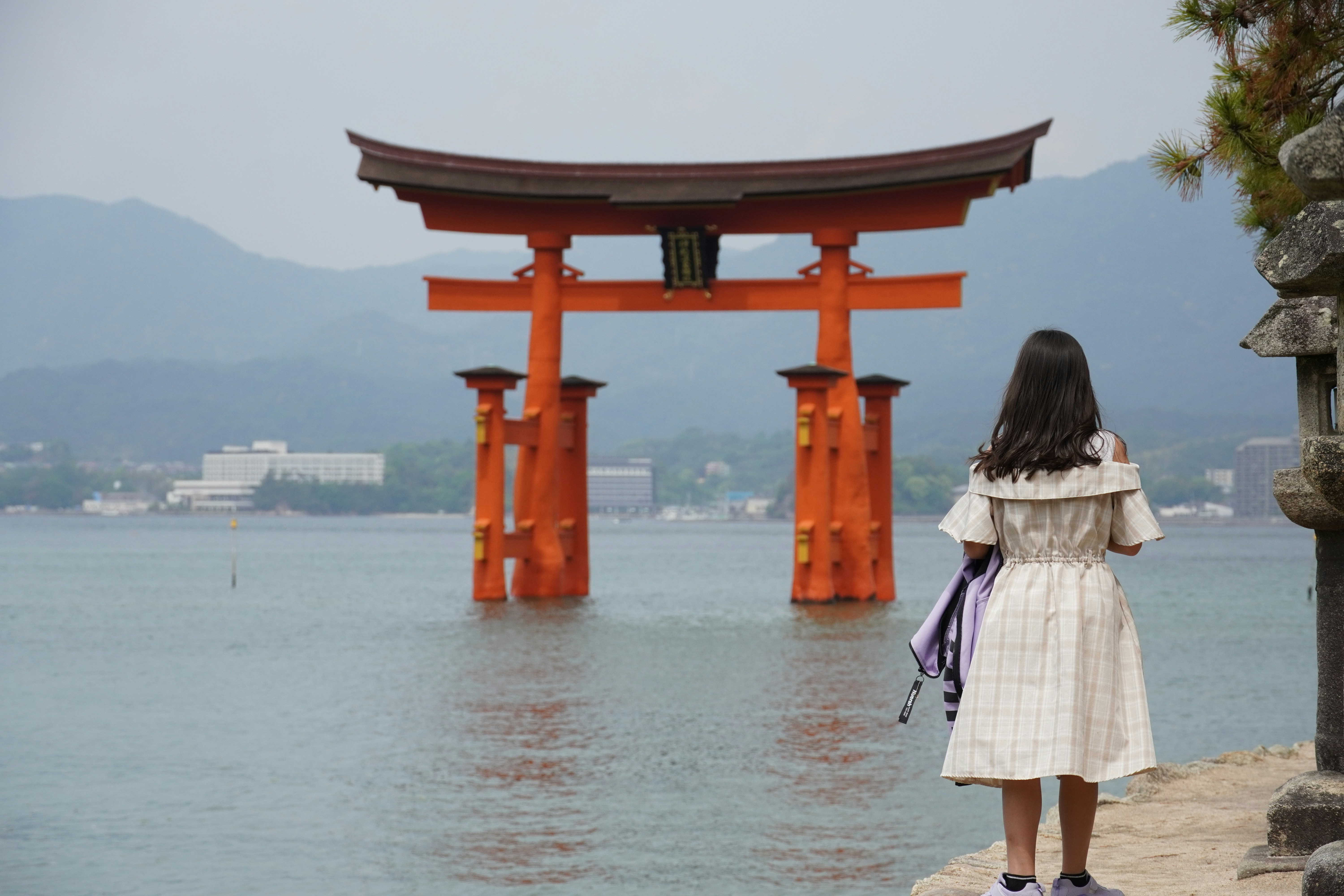 Person in a dress standing by the waterfront, gazing at the iconic red torii gate of Itsukushima Shrine.
