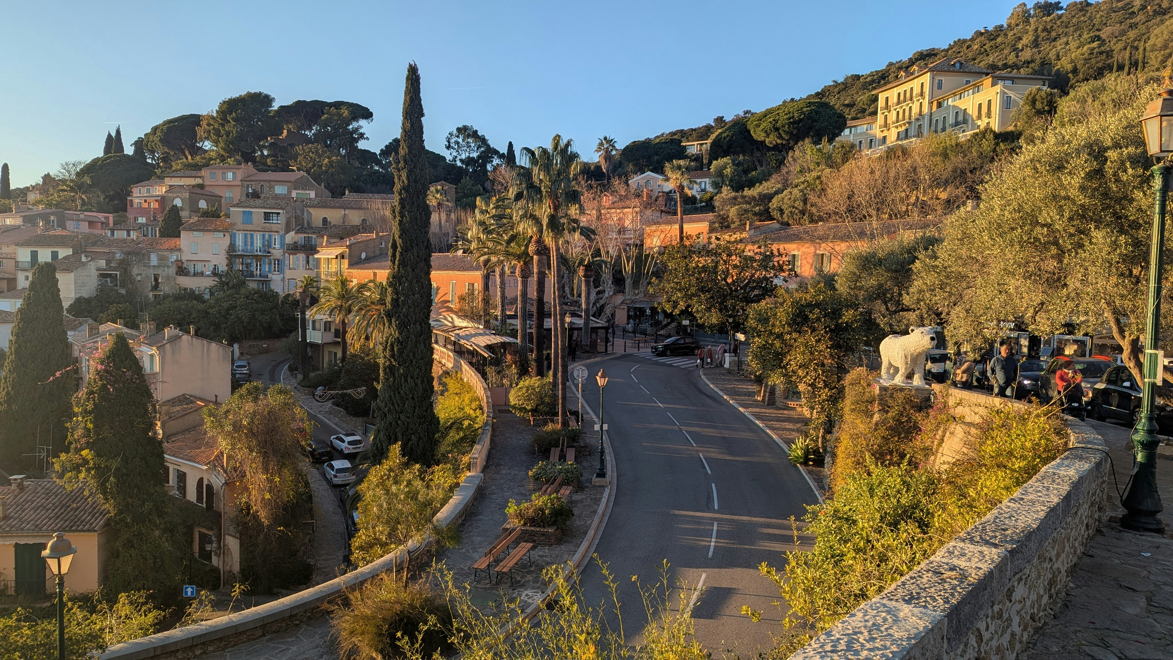 Curving road through a Mediterranean village bathed in warm sunset light, with cypress and palm trees lining the street.