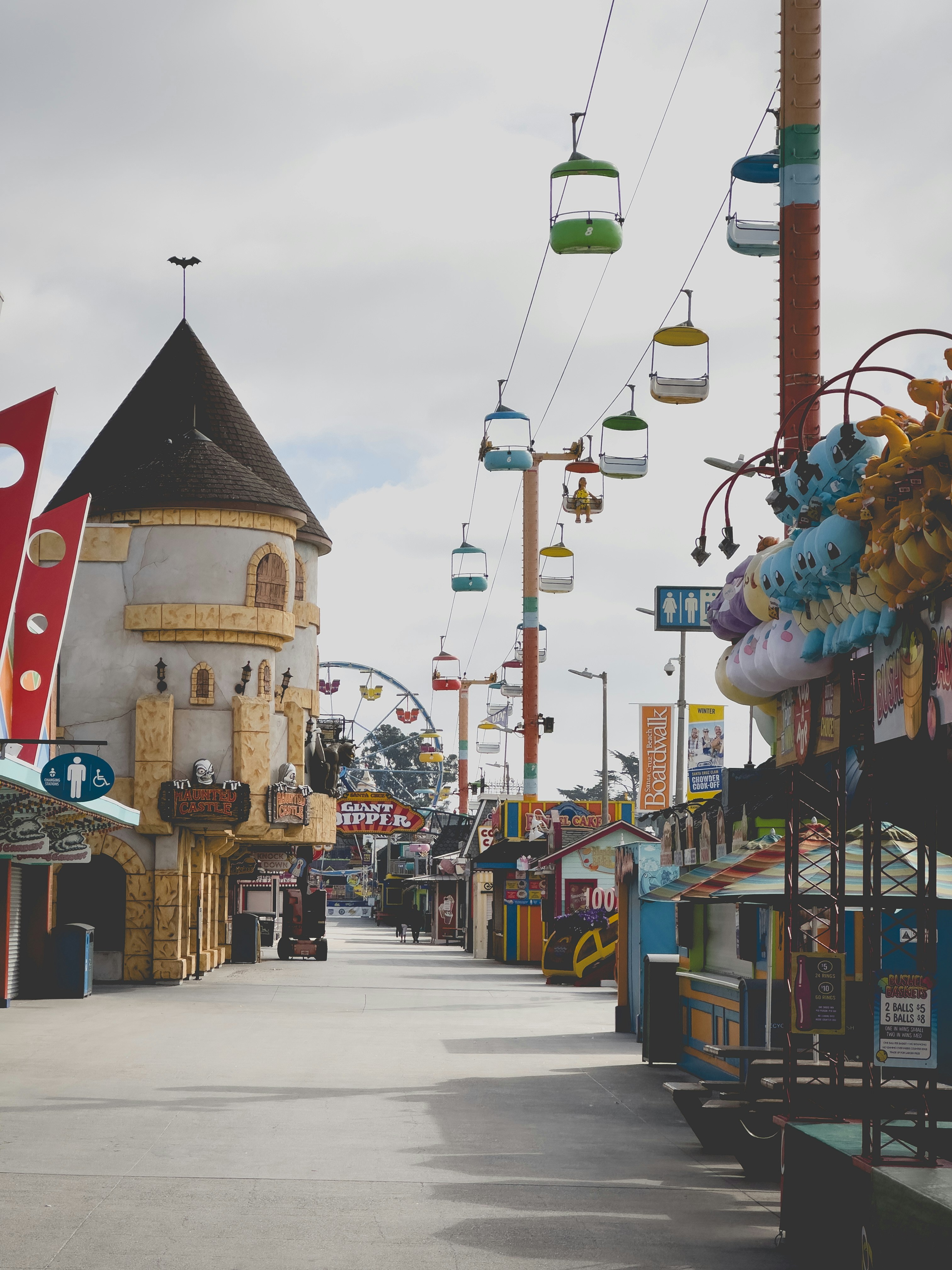 Quiet amusement park boardwalk with colorful cable cars and whimsical architecture under a cloudy sky.