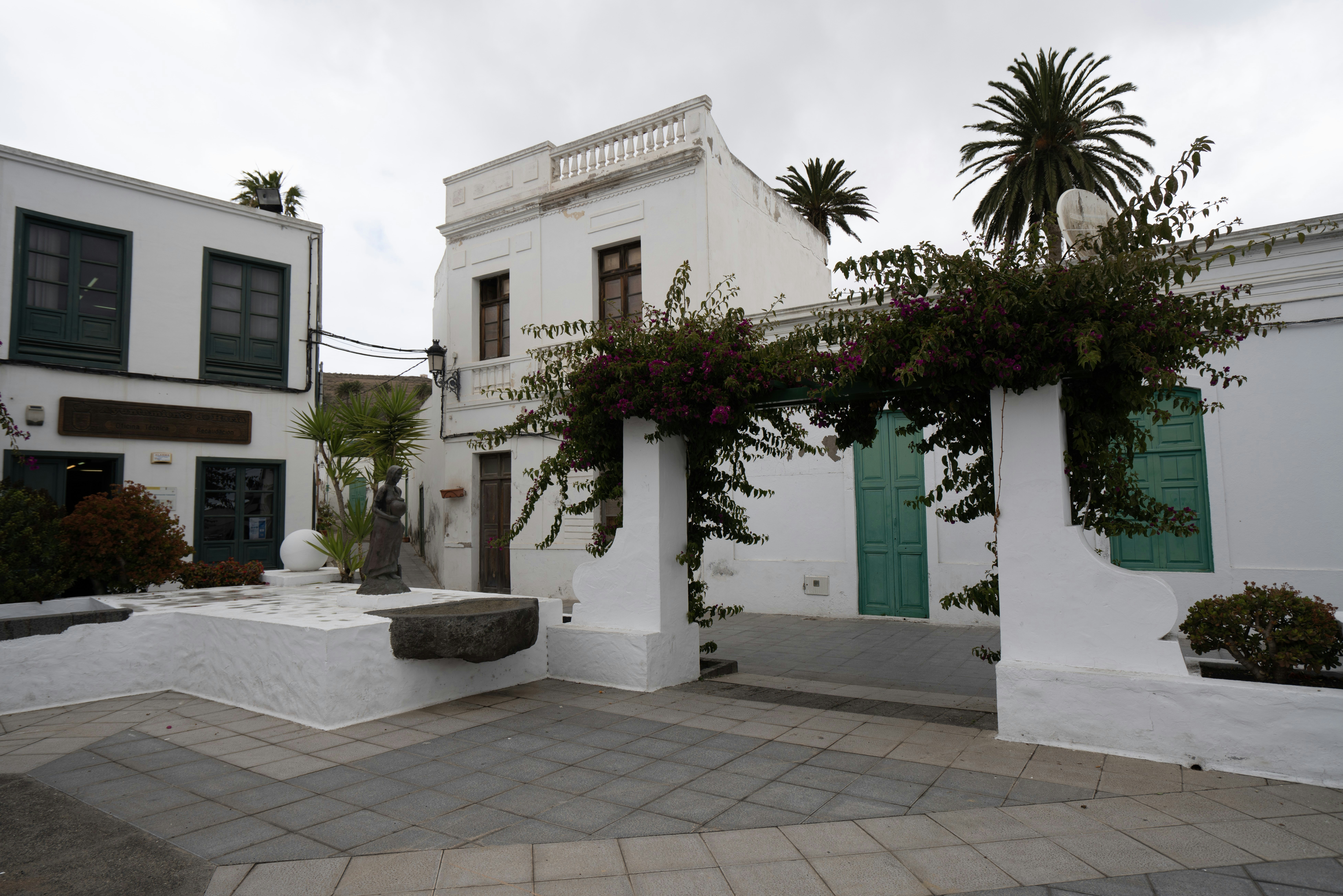 Whitewashed town square with green doors and blooming bougainvillea under a cloudy sky.