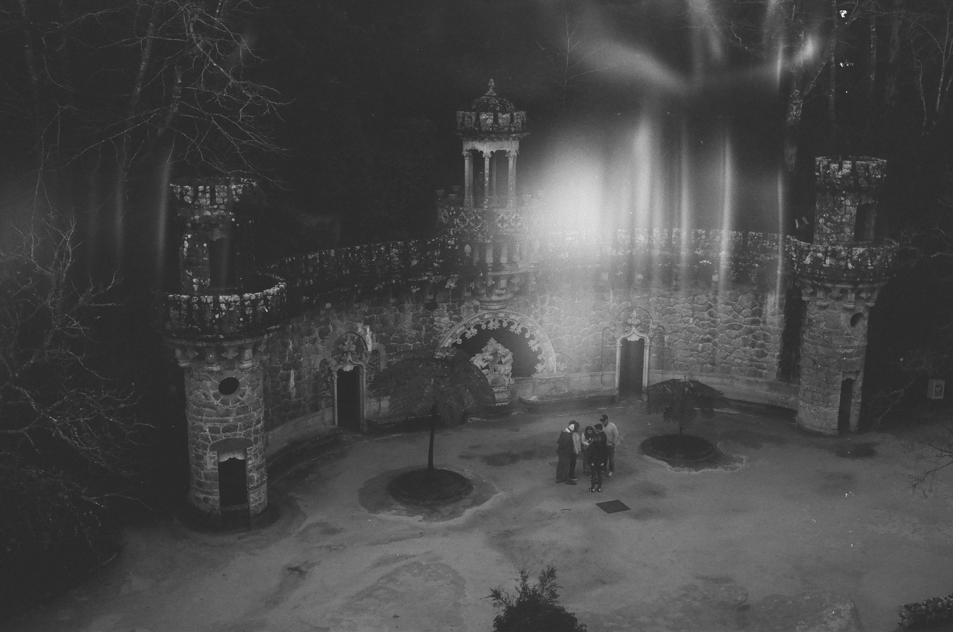 Black and white photograph of a small group standing before an ancient stone structure with turrets and archways under ethereal light streaks.