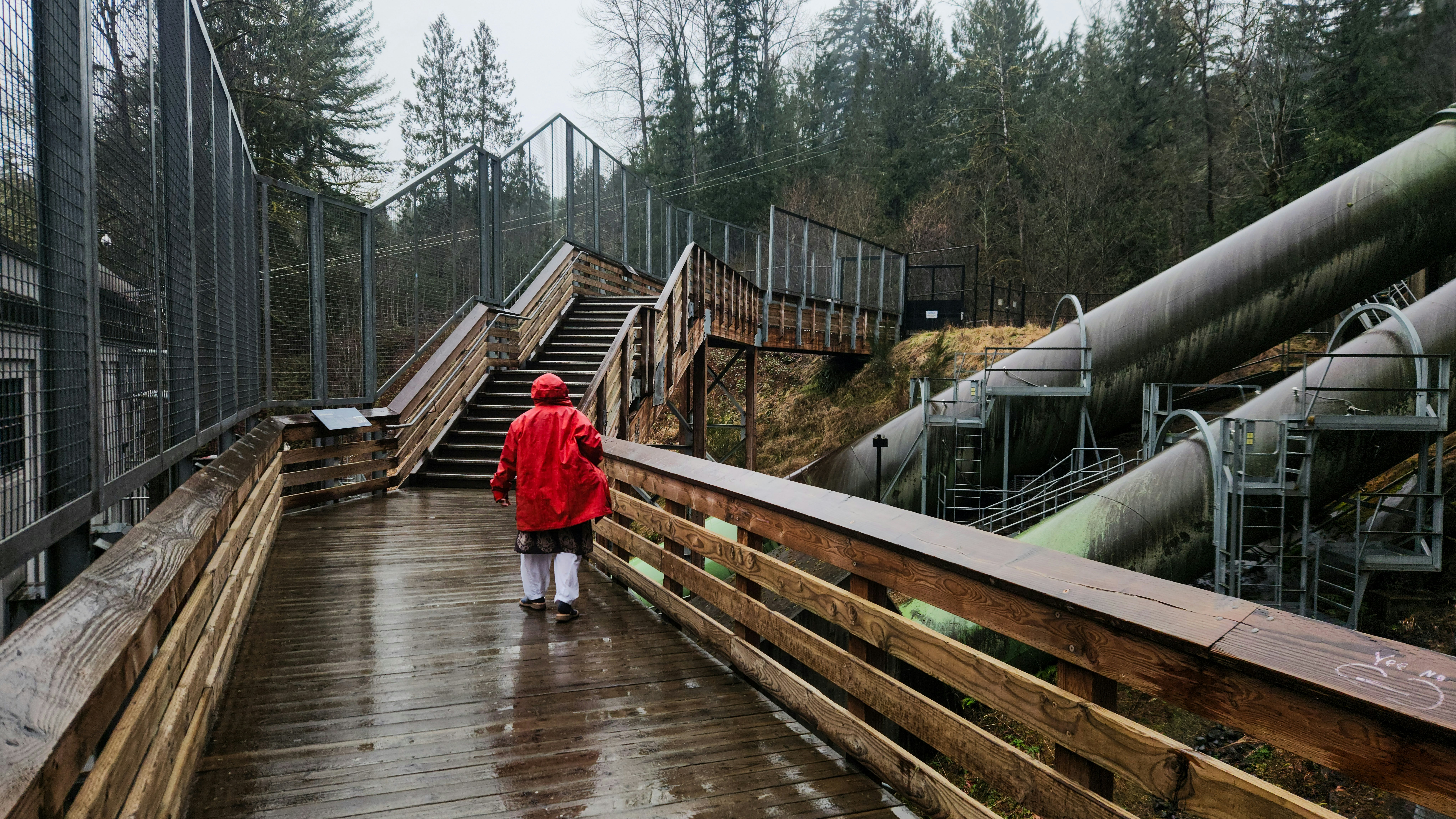 Visitor in red raincoat walking along a damp wooden boardwalk at Snoqualmie Falls Hydroelectric Facility amidst misty evergreen forest.