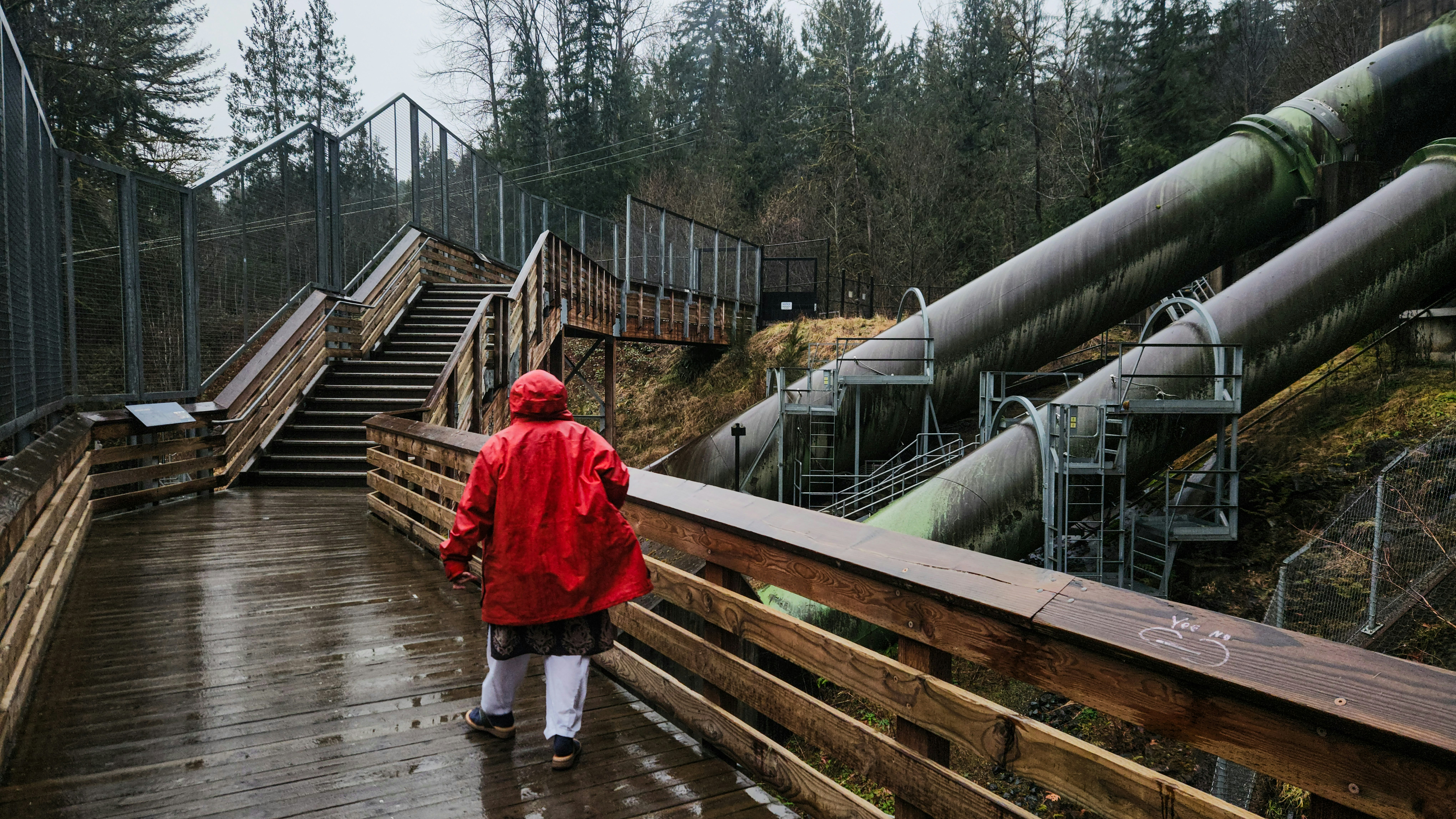 Person in a red raincoat on a wet wooden boardwalk near large green penstocks in a forested area.