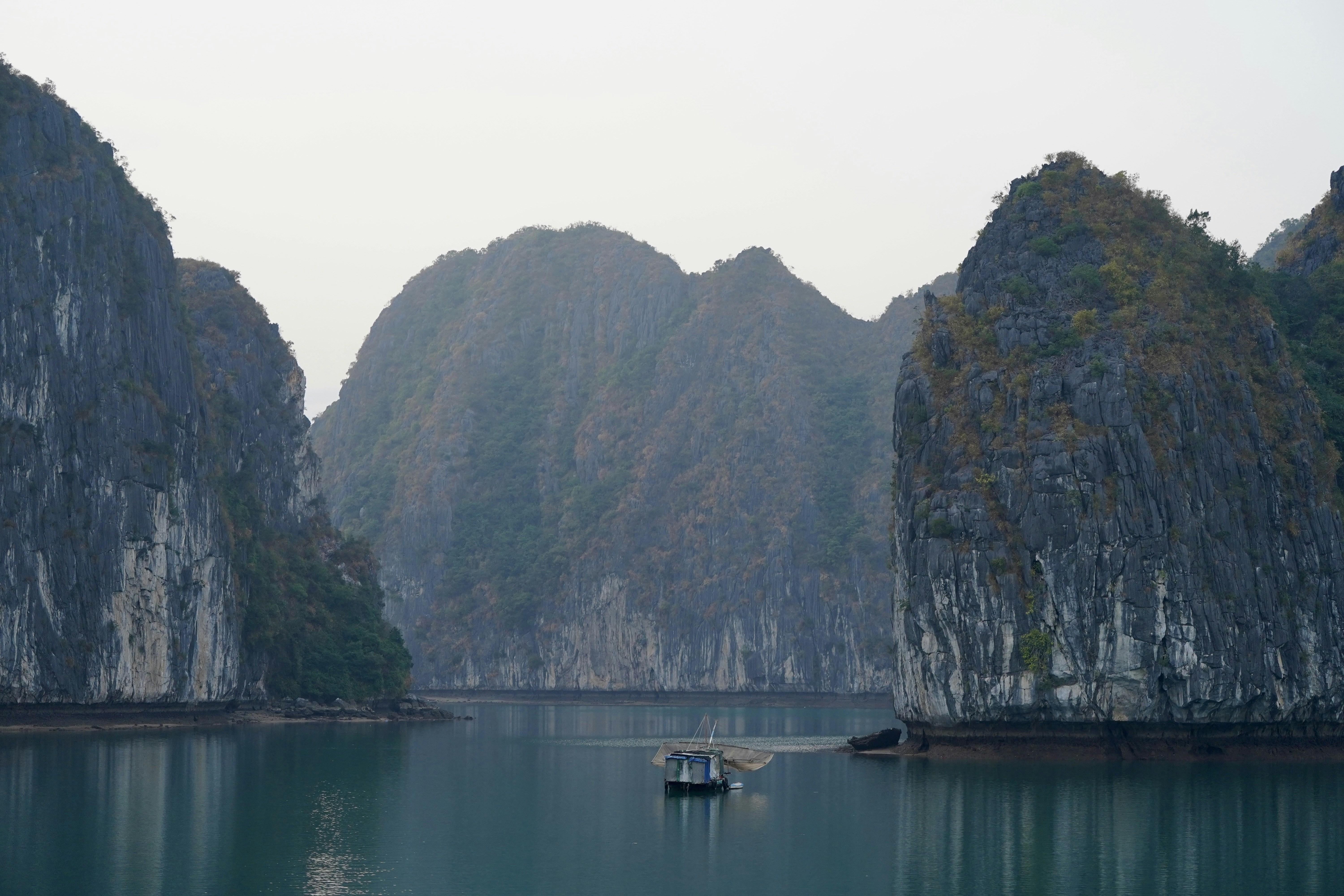 A boat navigates tranquil waters between towering limestone formations under an overcast sky.